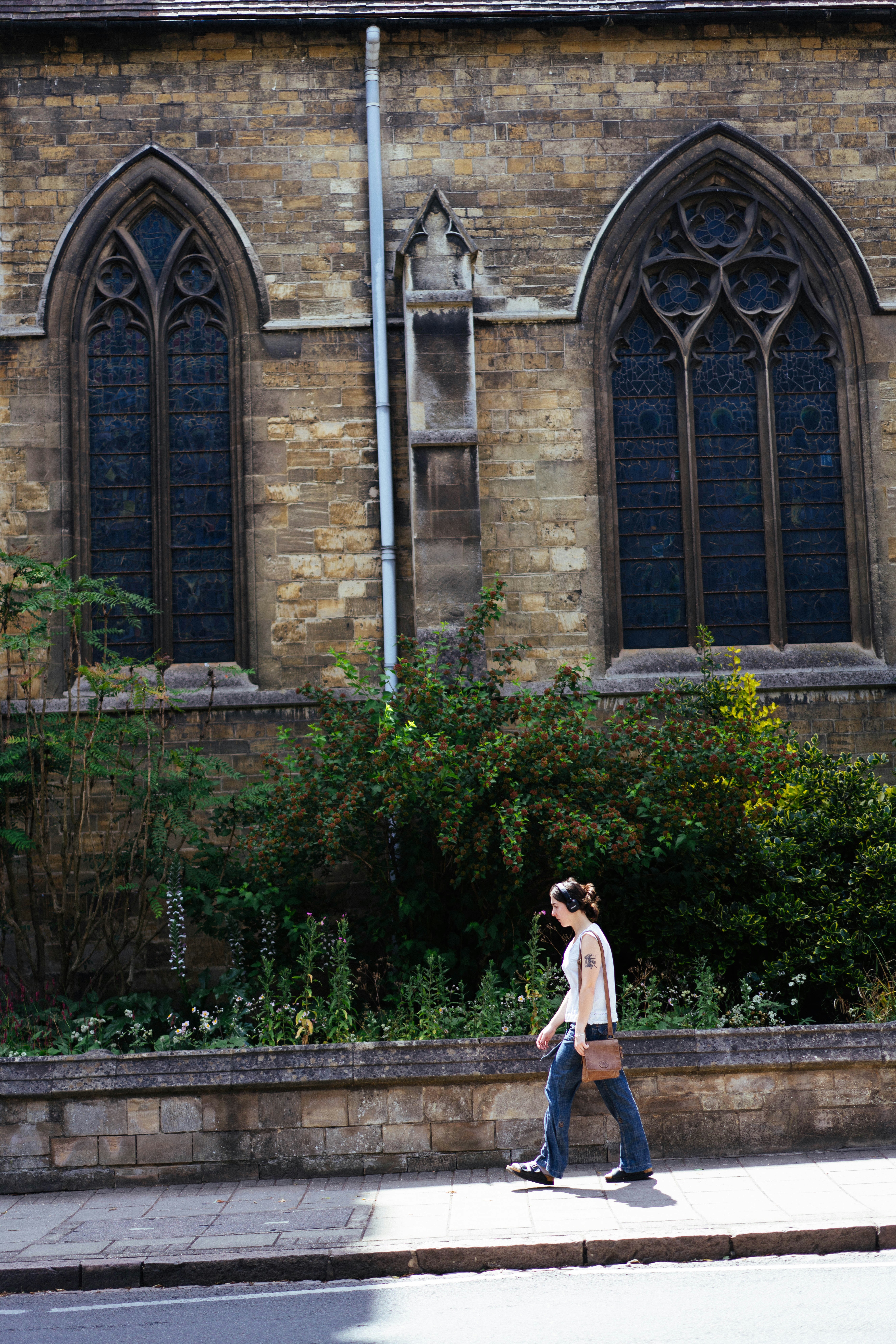 A person walks casually along a stone path beside a historic church, framed by lush greenery and intricate Gothic windows.