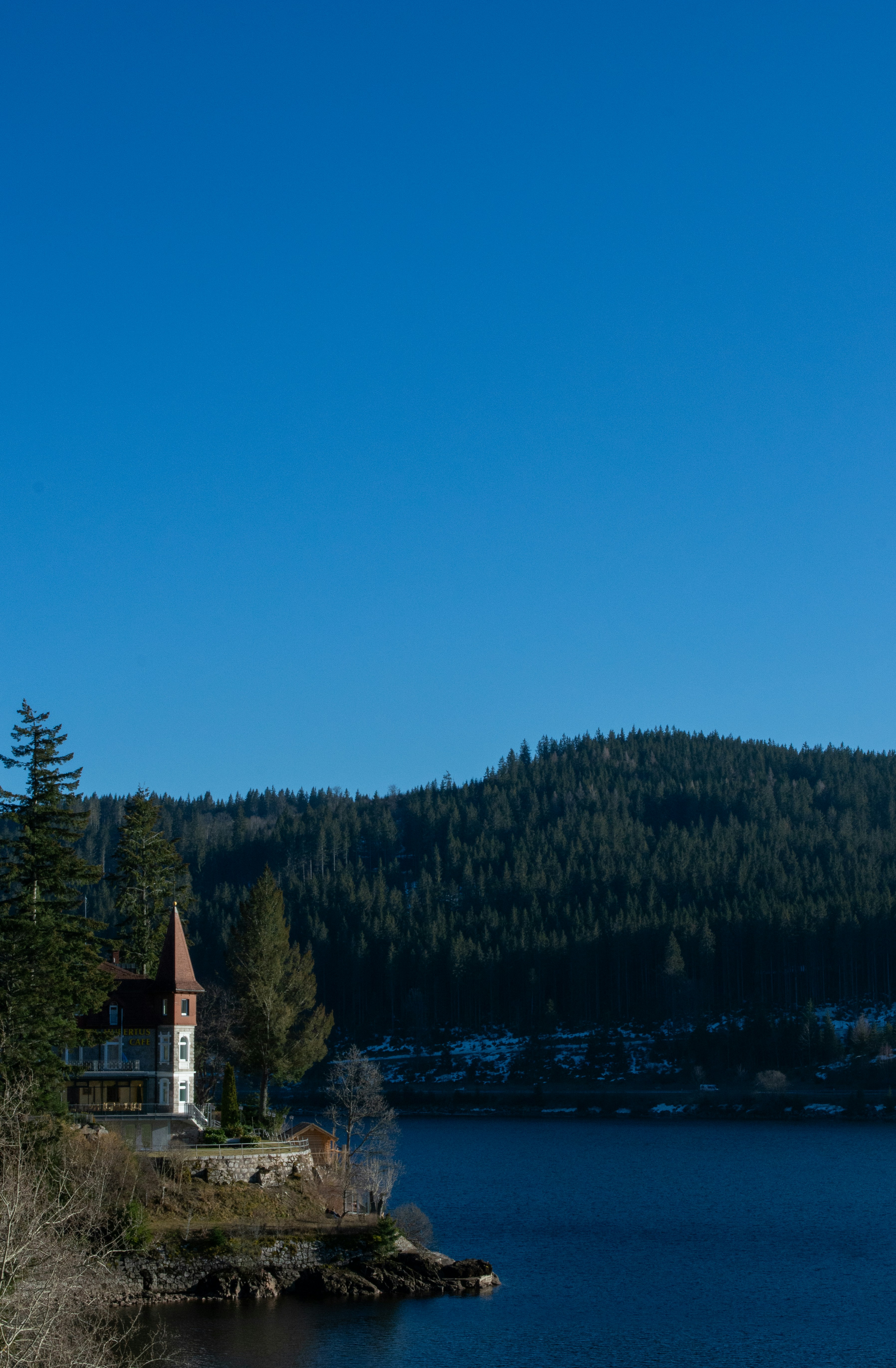 A charming turreted villa nestles along the shores of Schluchsee, overlooking calm blue waters and backed by dense, snow-dusted Black Forest hills. The tranquil setting captures the fairytale essence of the region, where architecture and alpine nature blend in quiet harmony. | A house overlooks the lake and forest.