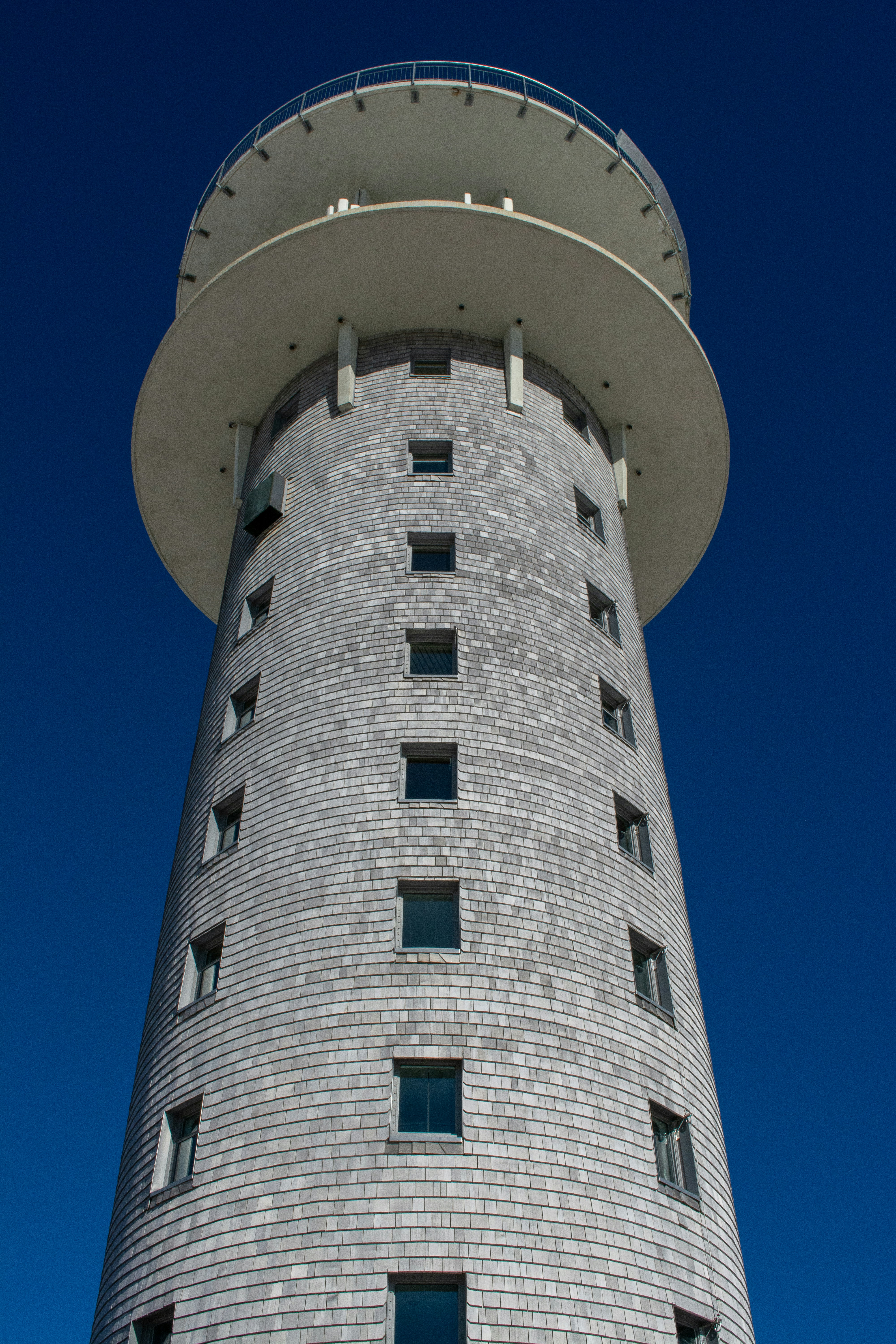 Soaring into a deep blue sky, the Feldberg Tower stands as a modern sentinel atop the Black Forest's highest peak. Its distinctive shingle-clad façade and circular observation deck echo both utility and design, offering panoramic views across the alpine horizon. | A tall tower is seen against a bright, blue sky.