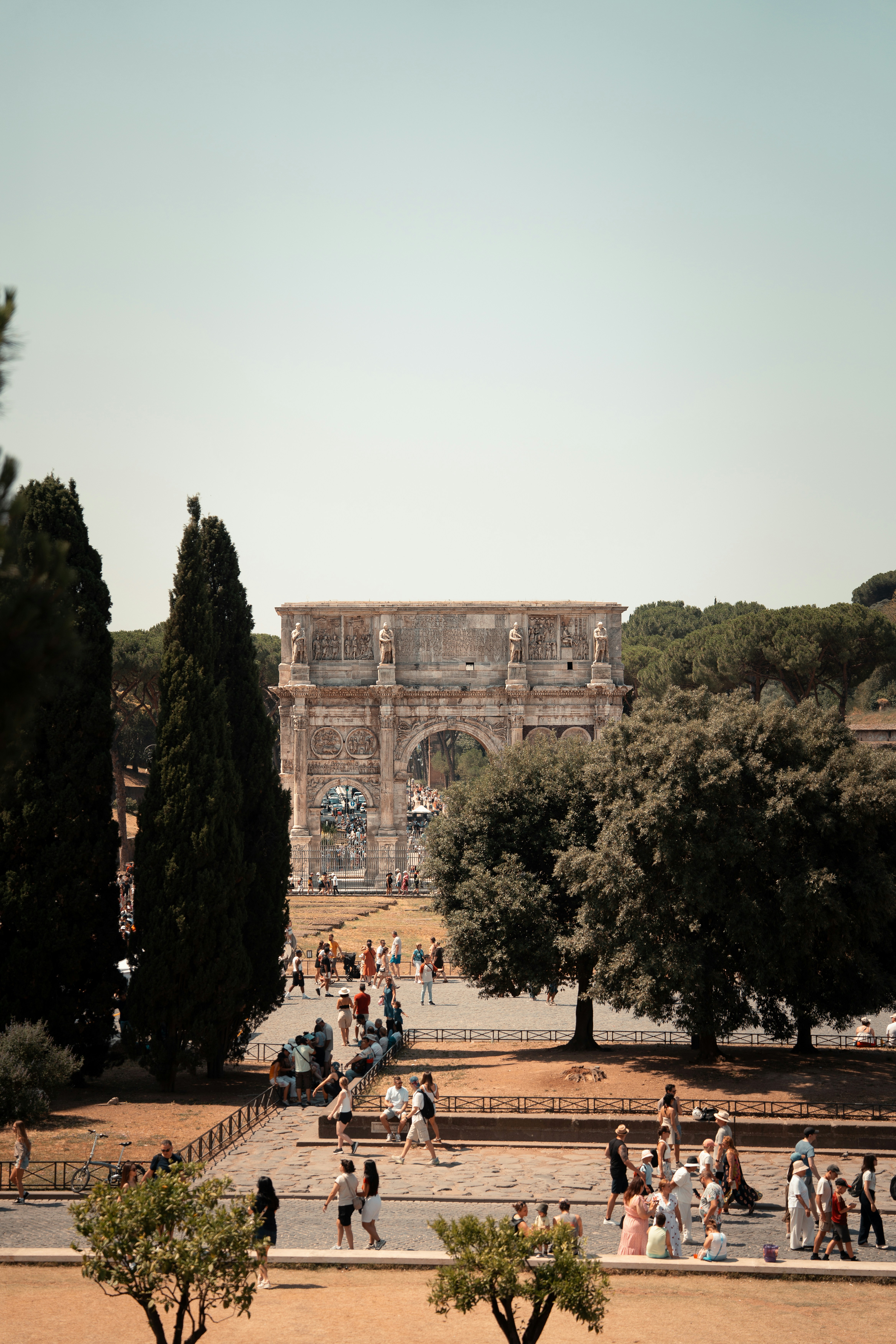 The Arch of Constantine stands majestically amidst a bustling crowd, framed by lush greenery and historical pathways.