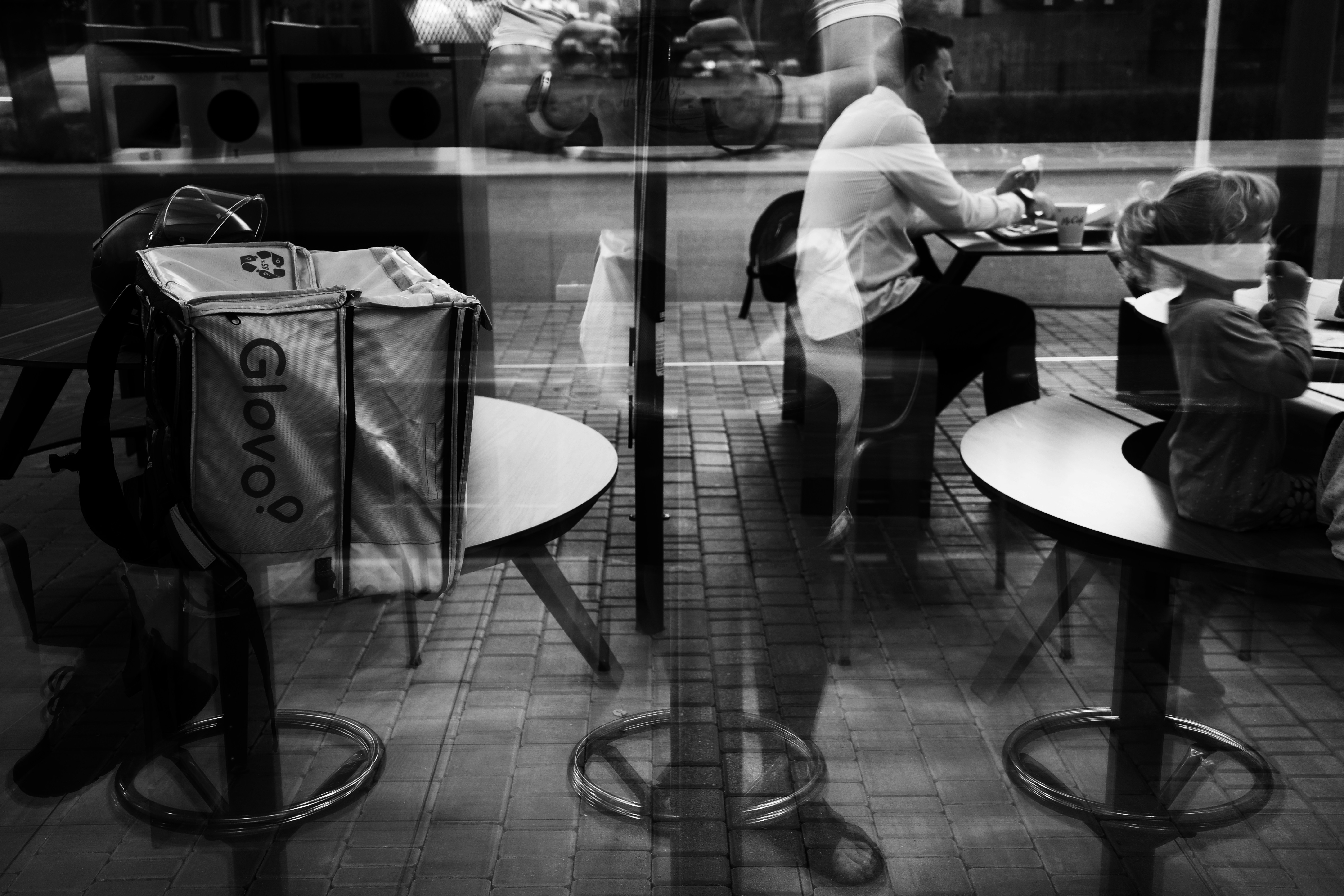 A black and white street scene captures reflections and activity through a window, blending interior and exterior elements. A distinctive Glovo delivery bag rests on a table in the foreground, while blurred figures, including an adult and a child, are seated at other tables within what appears to be a cafe or restaurant. The reflections on the glass create a layered, almost ghost-like effect, adding depth and intrigue to the urban tableau.