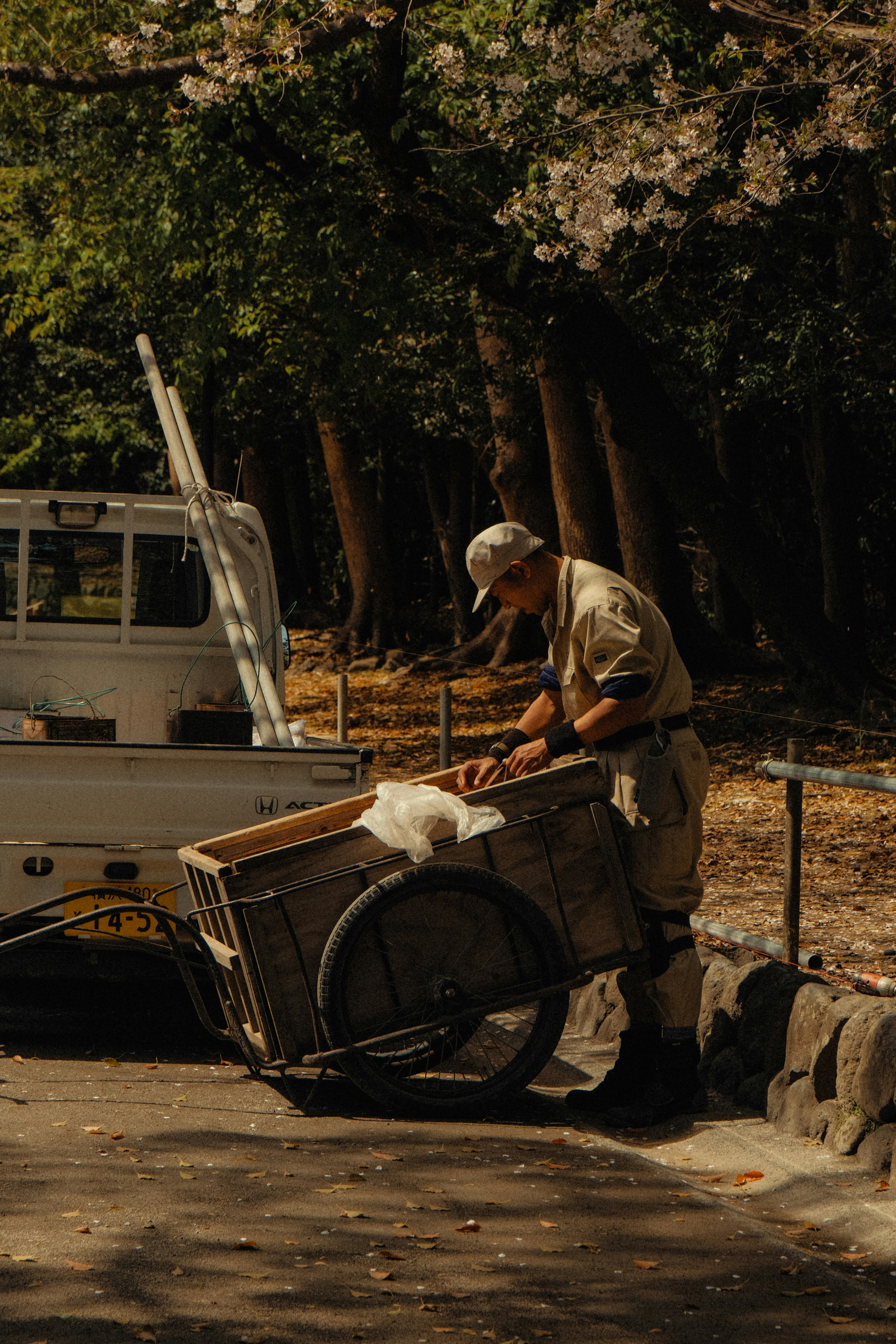 Dappled Duty: Park Worker Adjusts a Wooden Cart Under Leafy Canopy