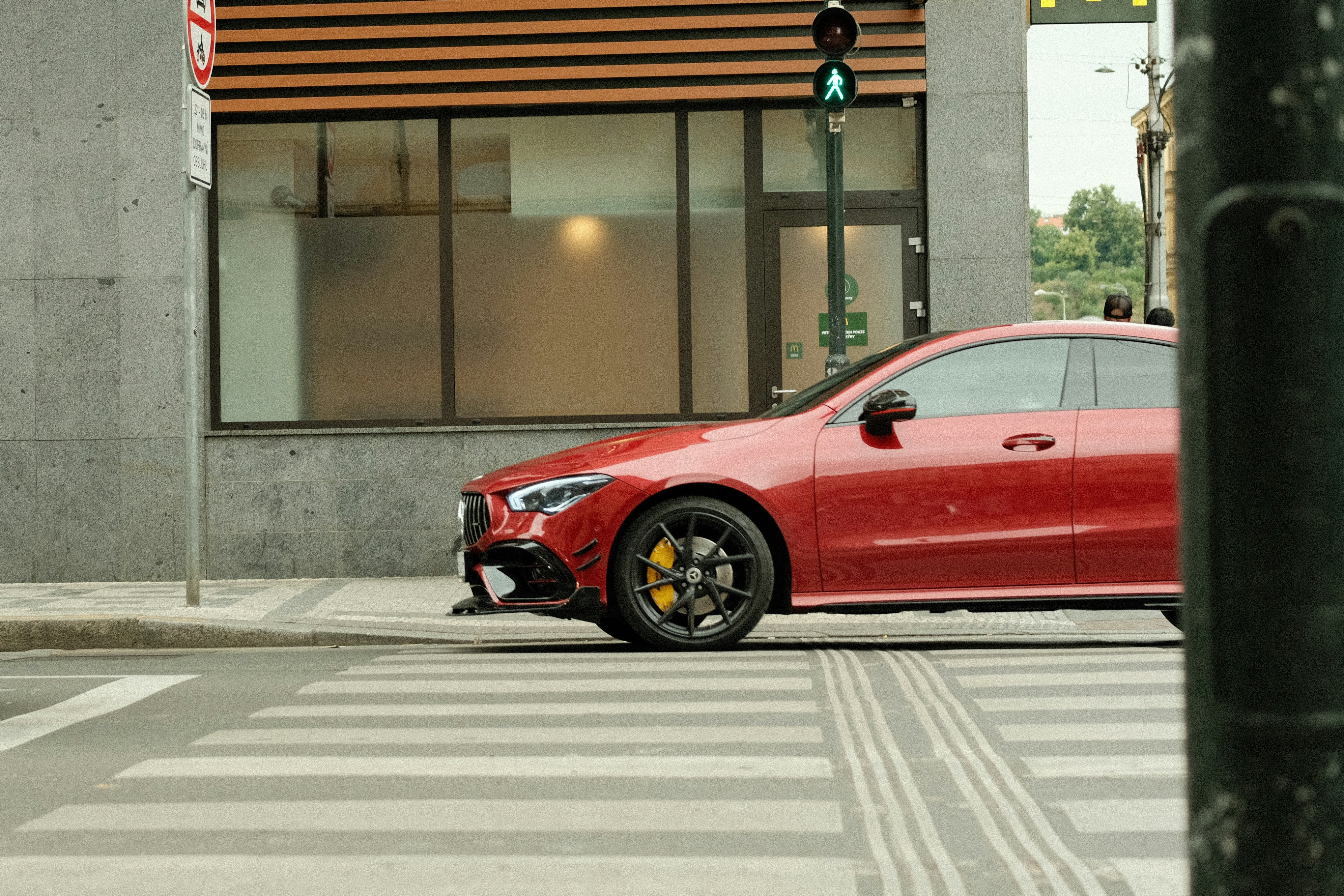 Red luxury car poised at a crosswalk in a modern urban setting, showcasing sleek design elements against a contemporary backdrop.