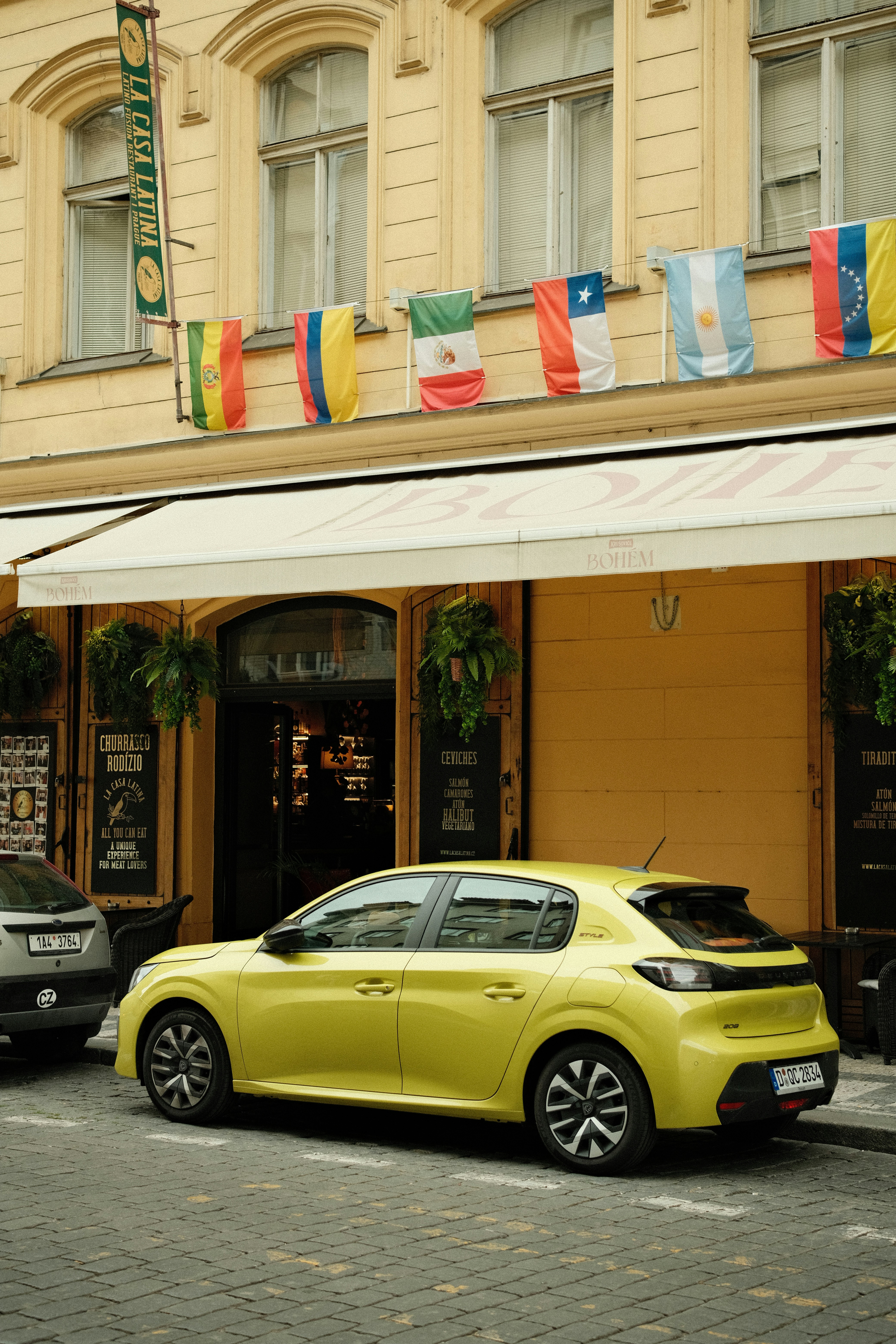 Peugeot 208. A yellow car is parked beside a restaurant featuring several international flags. The restaurant has a covered entrance and greenery in planters. | A yellow car parks outside a european building.