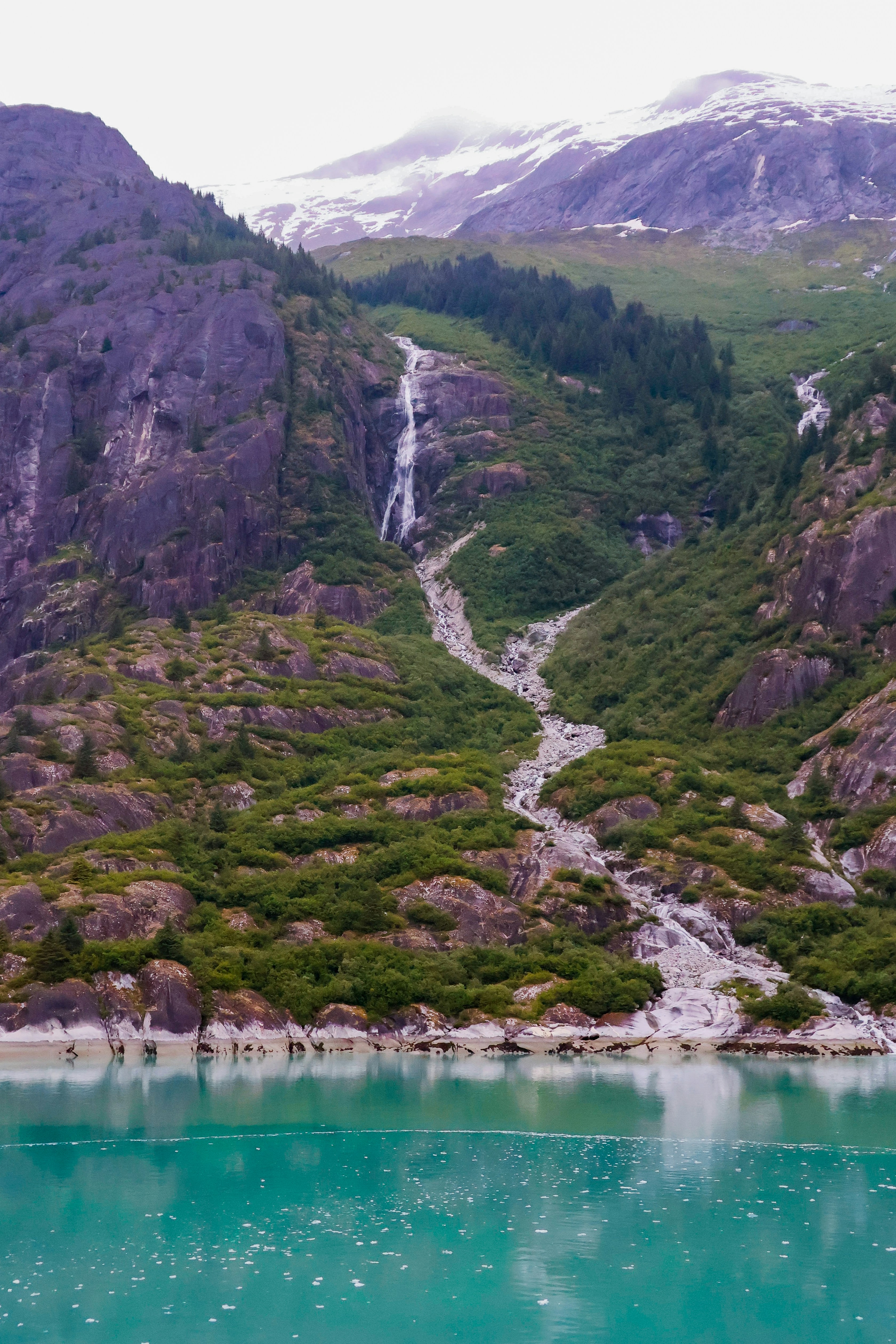 A tranquil mountain landscape featuring a cascading waterfall flowing down rocky terrain into a turquoise glacial lake, surrounded by lush greenery.