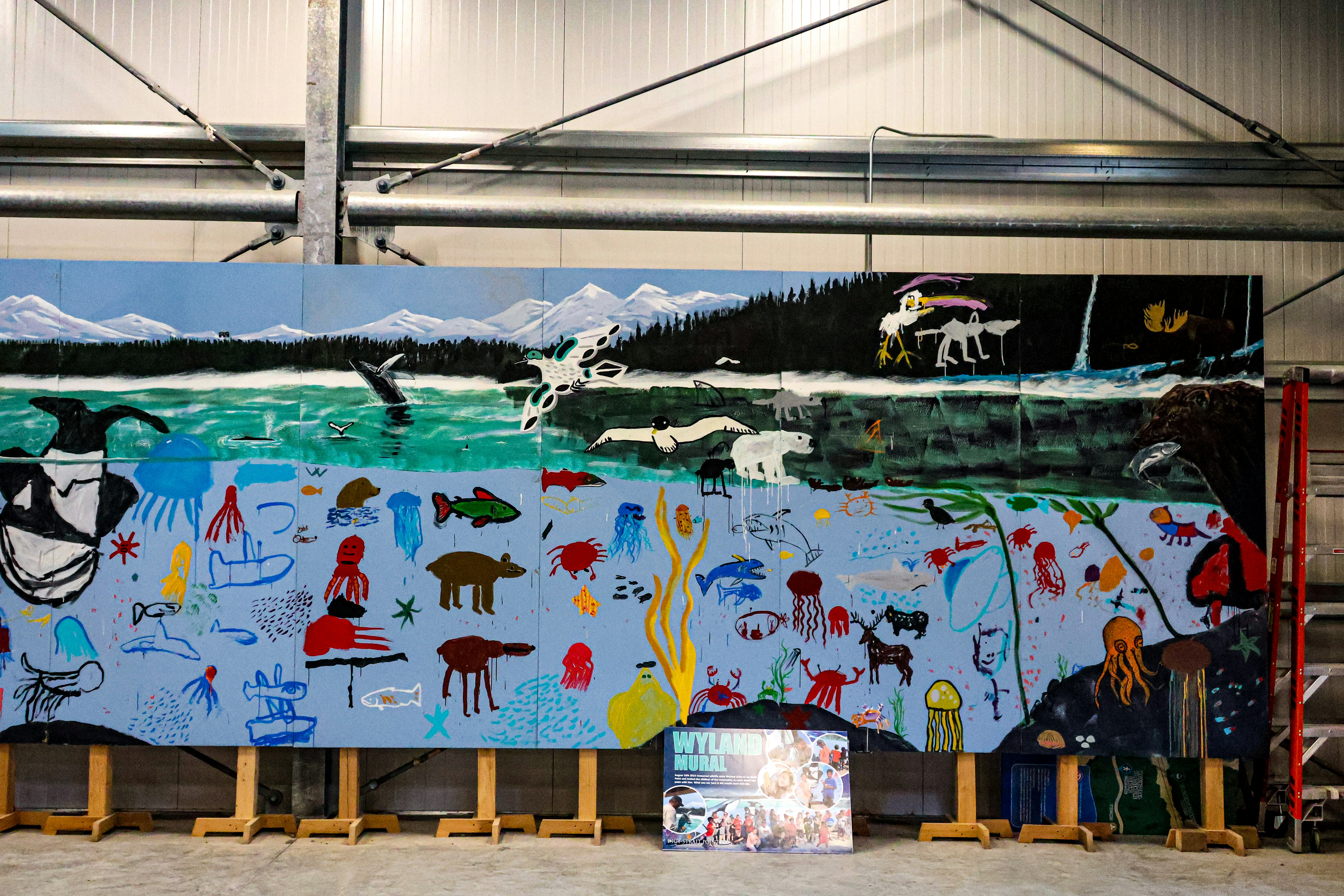 Vibrant mural depicting a lively underwater scene filled with various marine life and mythical creatures, set against a backdrop of mountains and a serene lake. A small poster is displayed at the bottom.