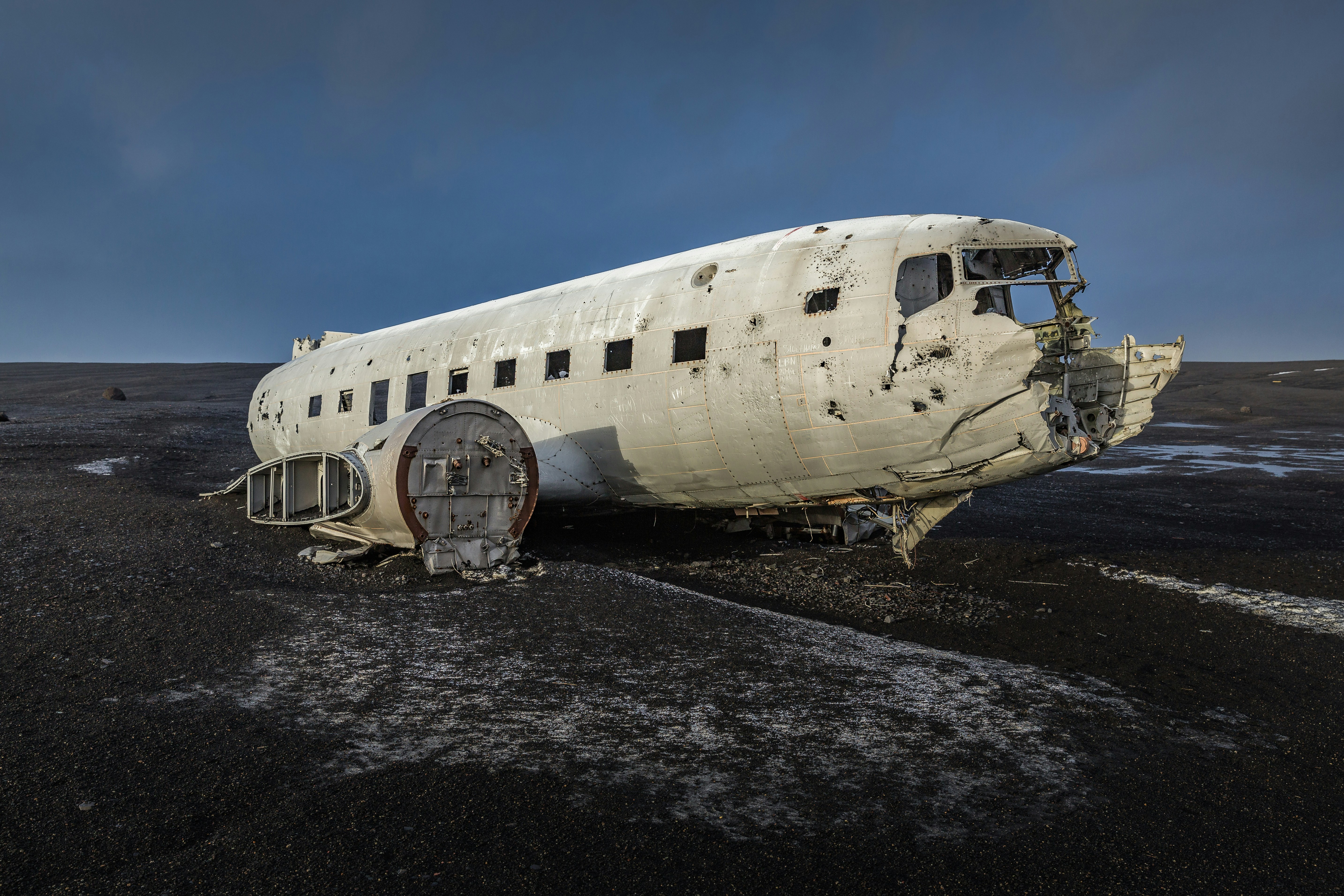 Abandoned airplane fuselage resting on a desolate black sand landscape, showcasing its weathered exterior against a moody sky.