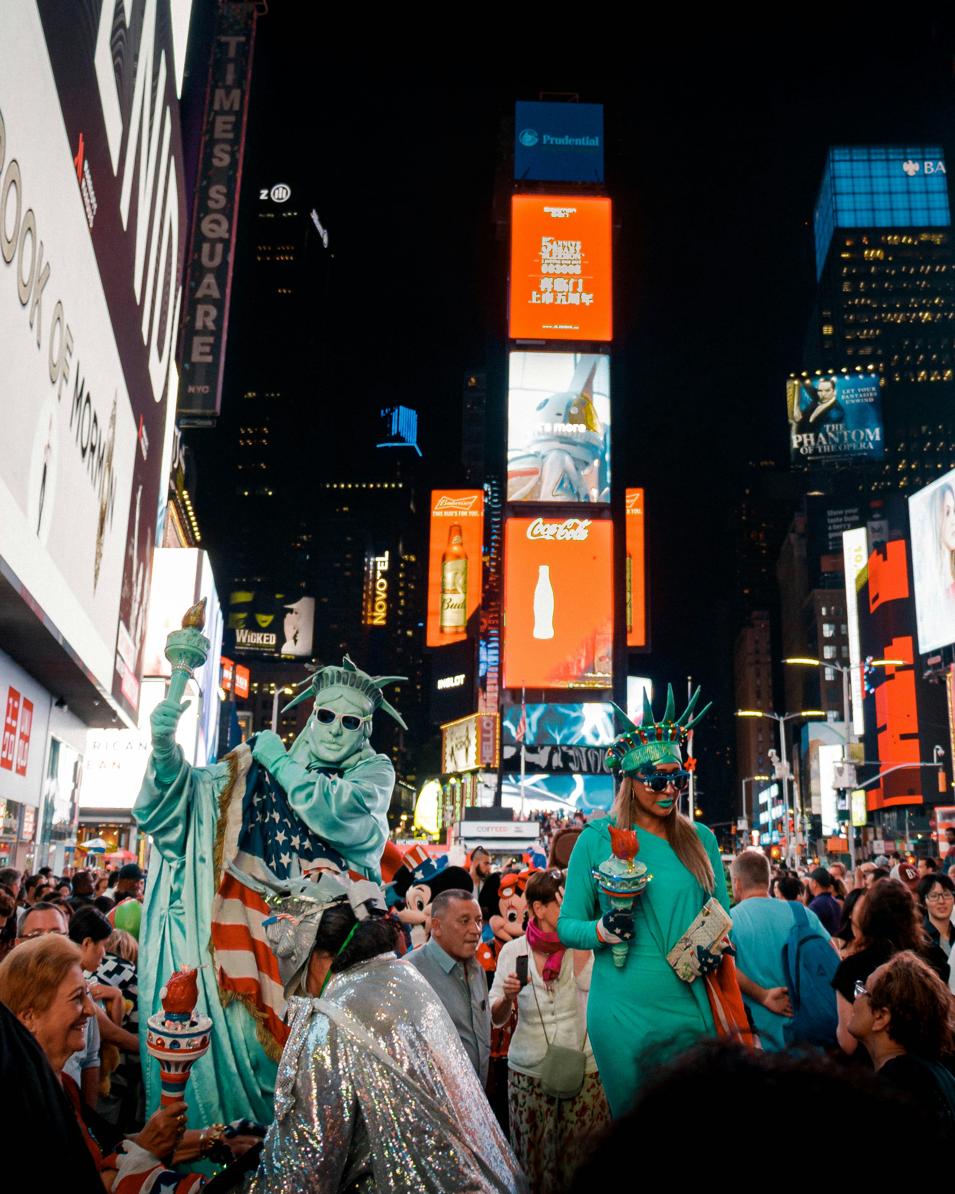 Two street performers dressed as the Statue of Liberty in Times Square, New York City, surrounded by a nighttime crowd. Urban street performance in a tourist hotspot. | Times square at night, featuring statue of liberty impersonators.