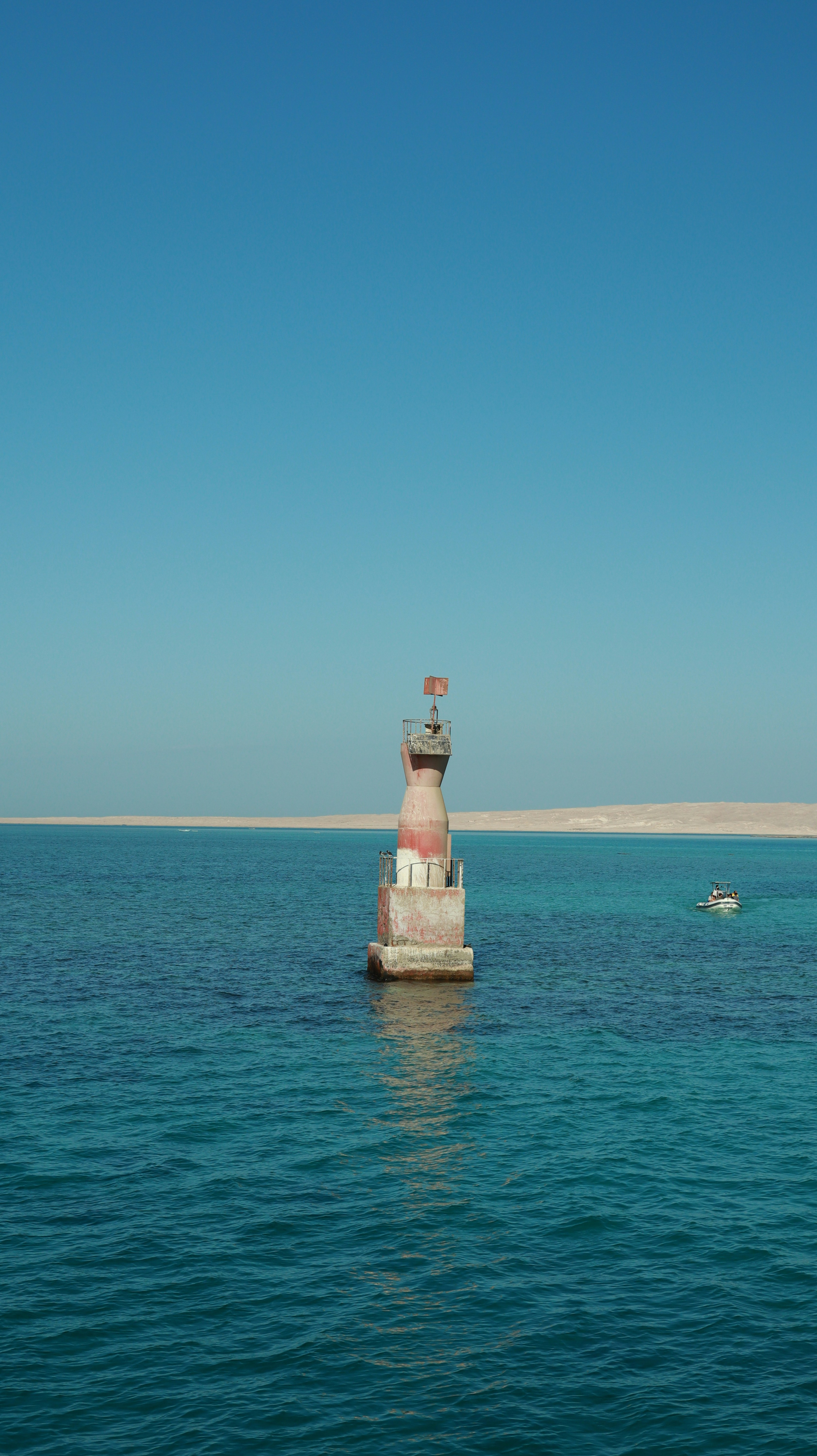 Weathered lighthouse standing tall in calm turquoise waters against a clear blue sky. A small boat glides by in the distance.