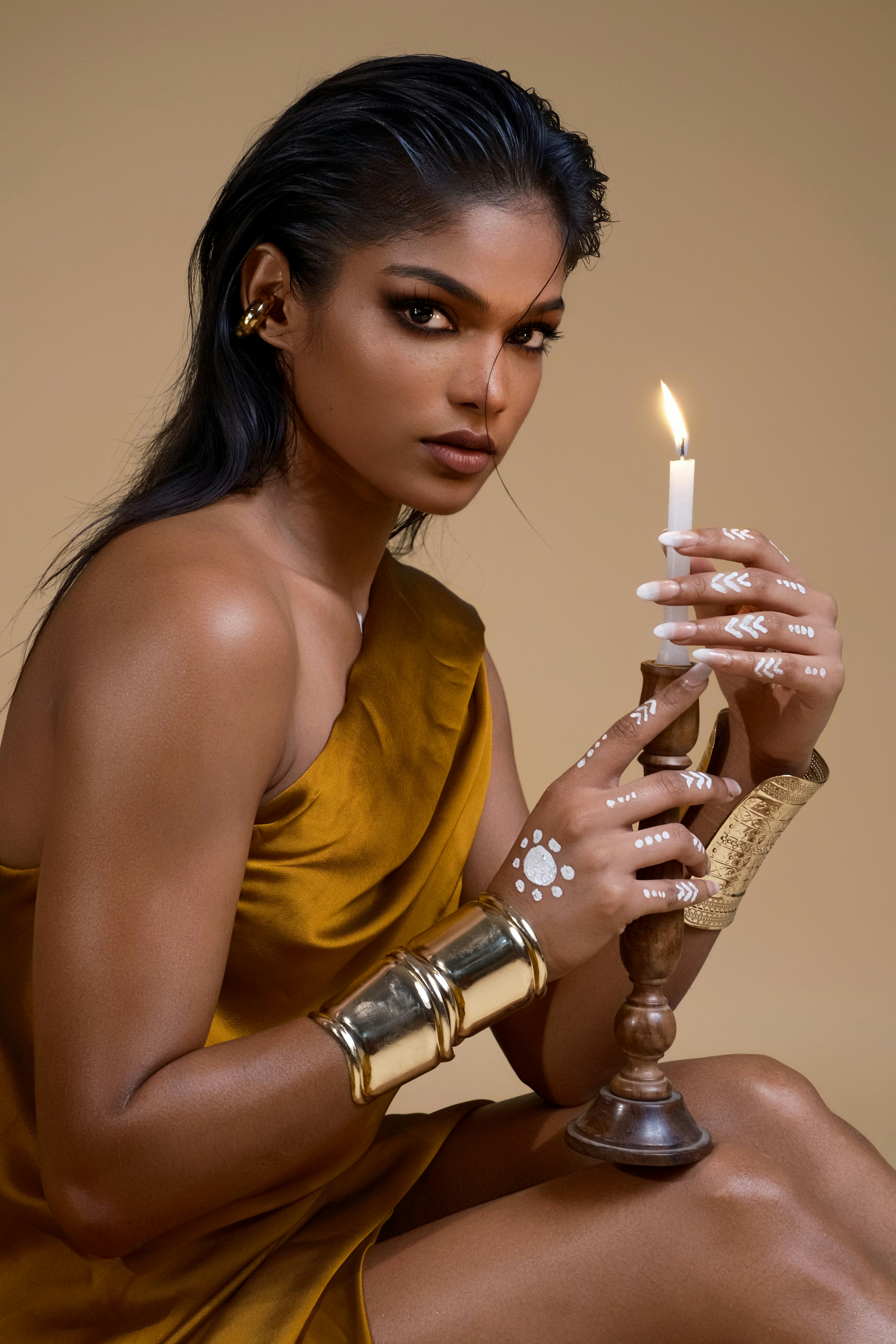 Woman in a golden drape holding a lit candle, adorned with intricate jewelry and body art against a warm beige backdrop.