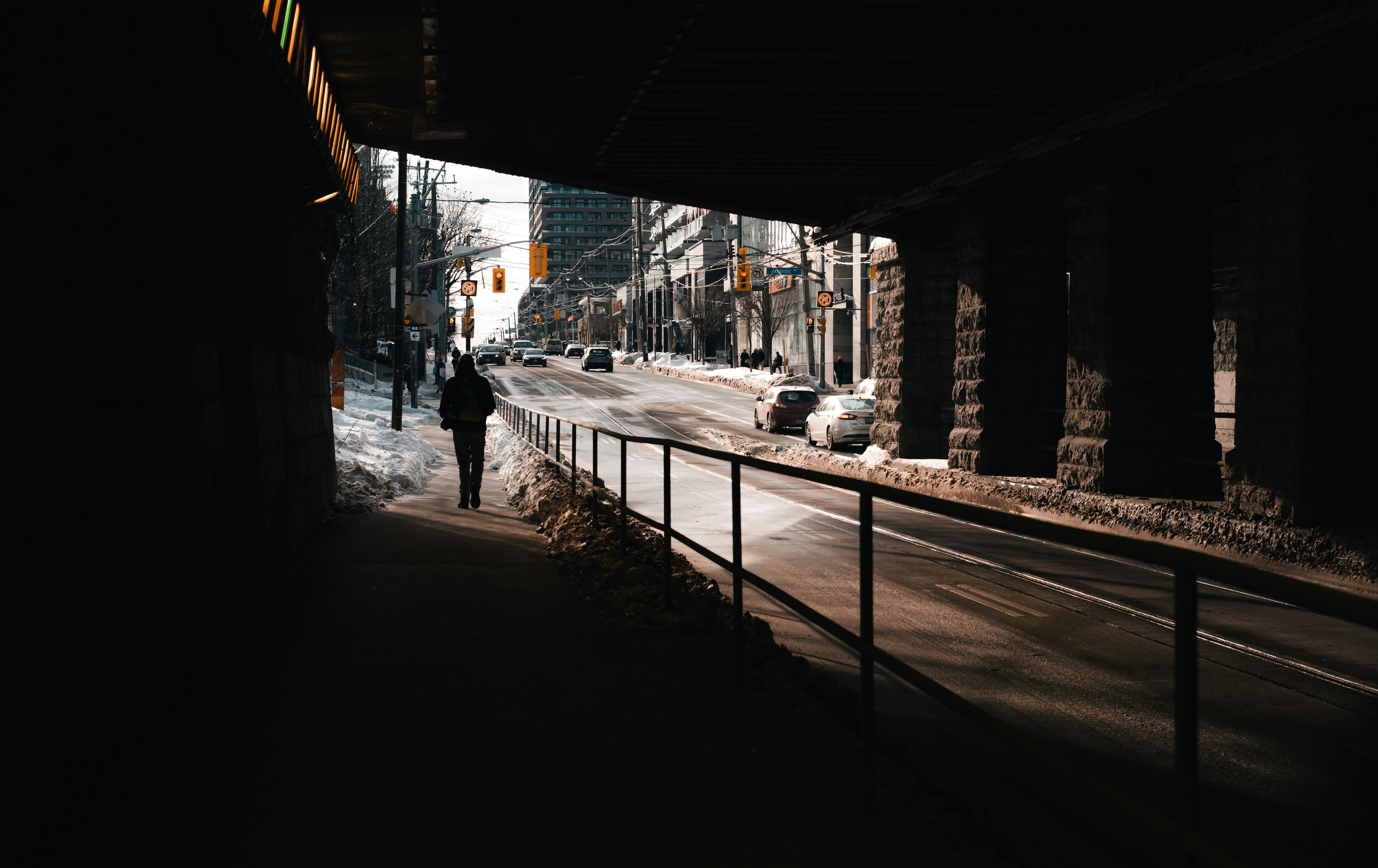 A lone figure walks along a snow-dusted sidewalk under a bridge, illuminated by soft city lights. The scene captures the contrast between shadow and light in an urban setting.