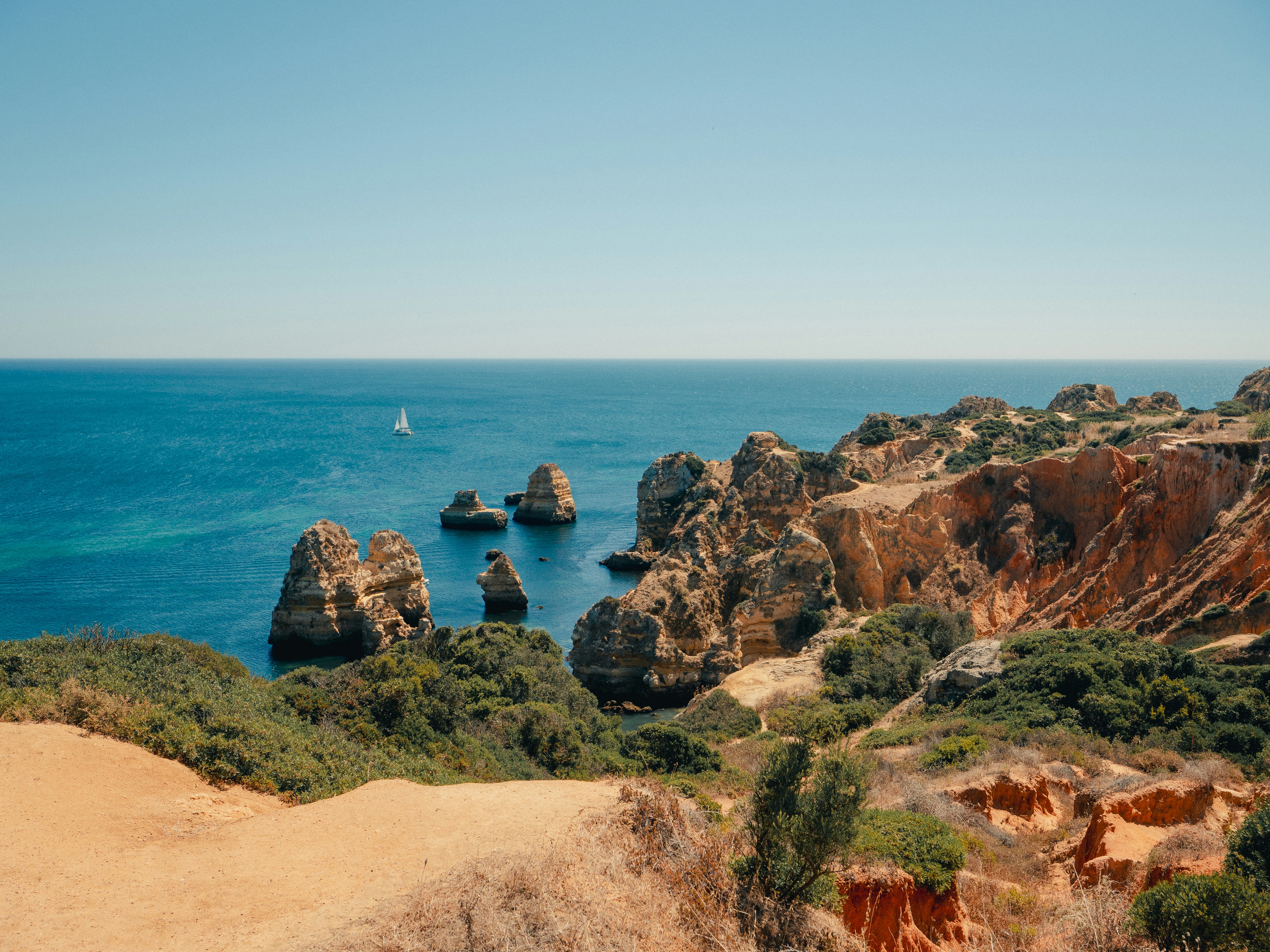 Rock formations rise majestically from the azure waters, with a sailboat gliding in the distance under a clear sky.