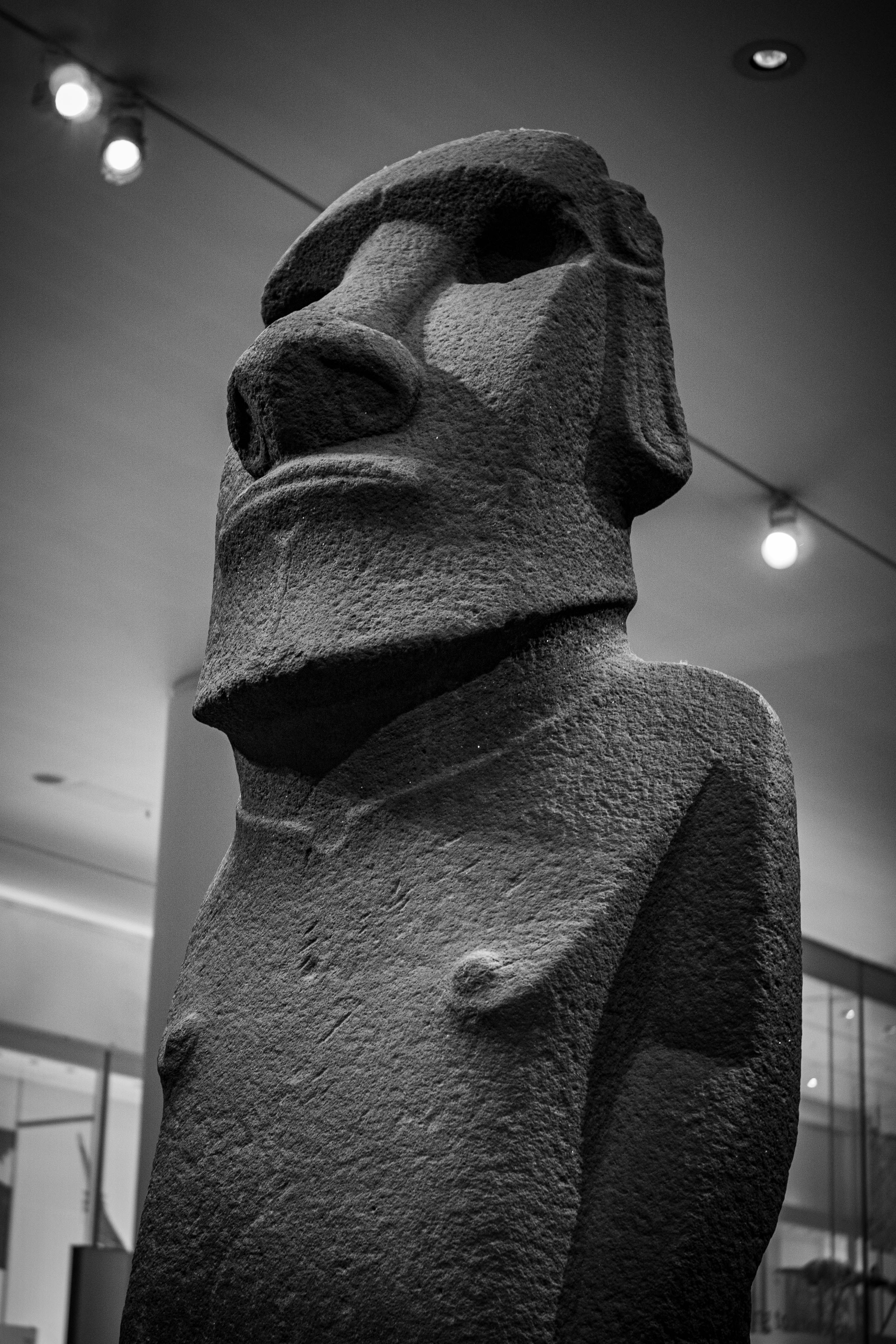 A monumental stone statue stands in a museum, showcasing intricate facial features and a stoic expression. The monochrome palette enhances its historical significance.