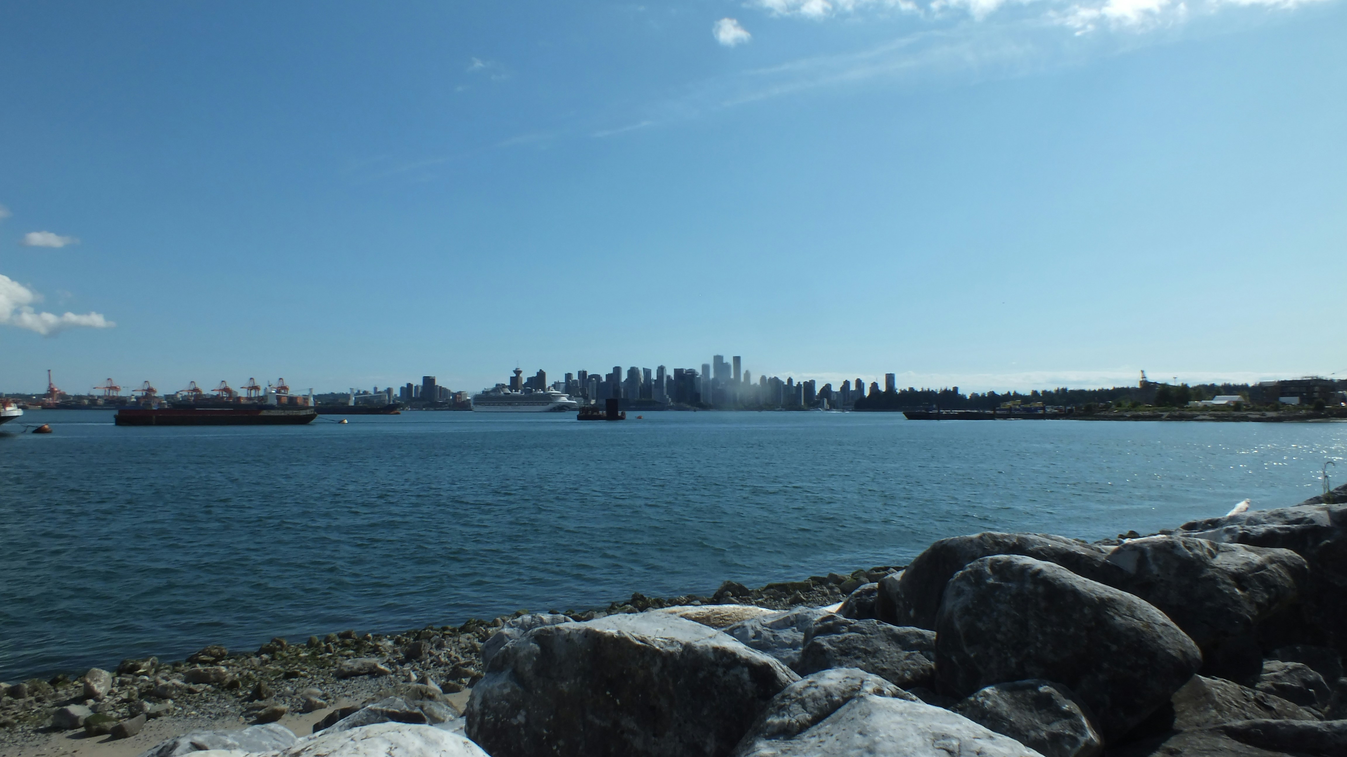 A serene view of a city skyline across the water, framed by rocky shoreline and anchored ships. The clear blue sky enhances the urban landscape.