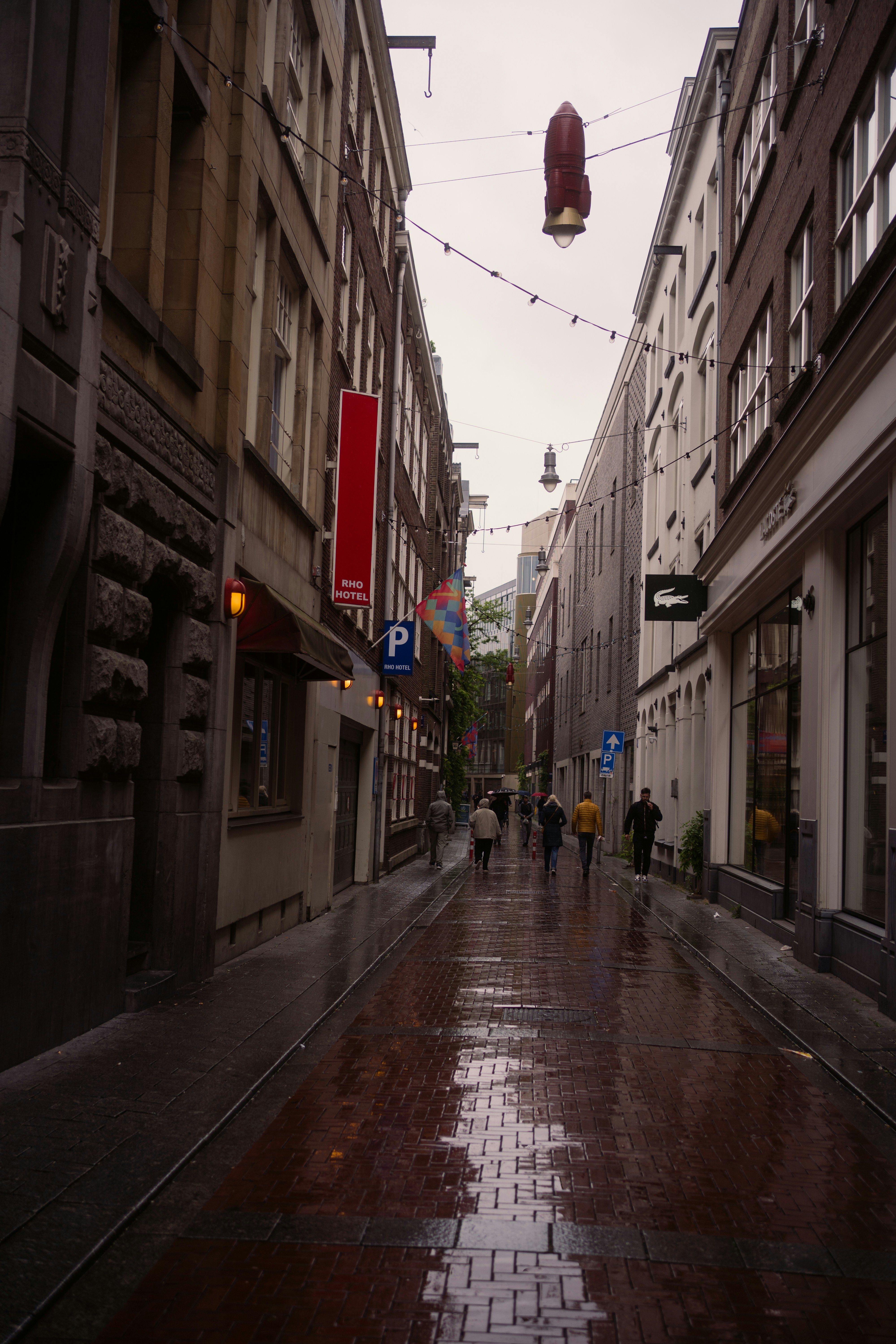 A narrow, cobblestone alleyway glistens after rain, with pedestrians casually walking under overcast skies, colorful flags and lights adding vibrancy.