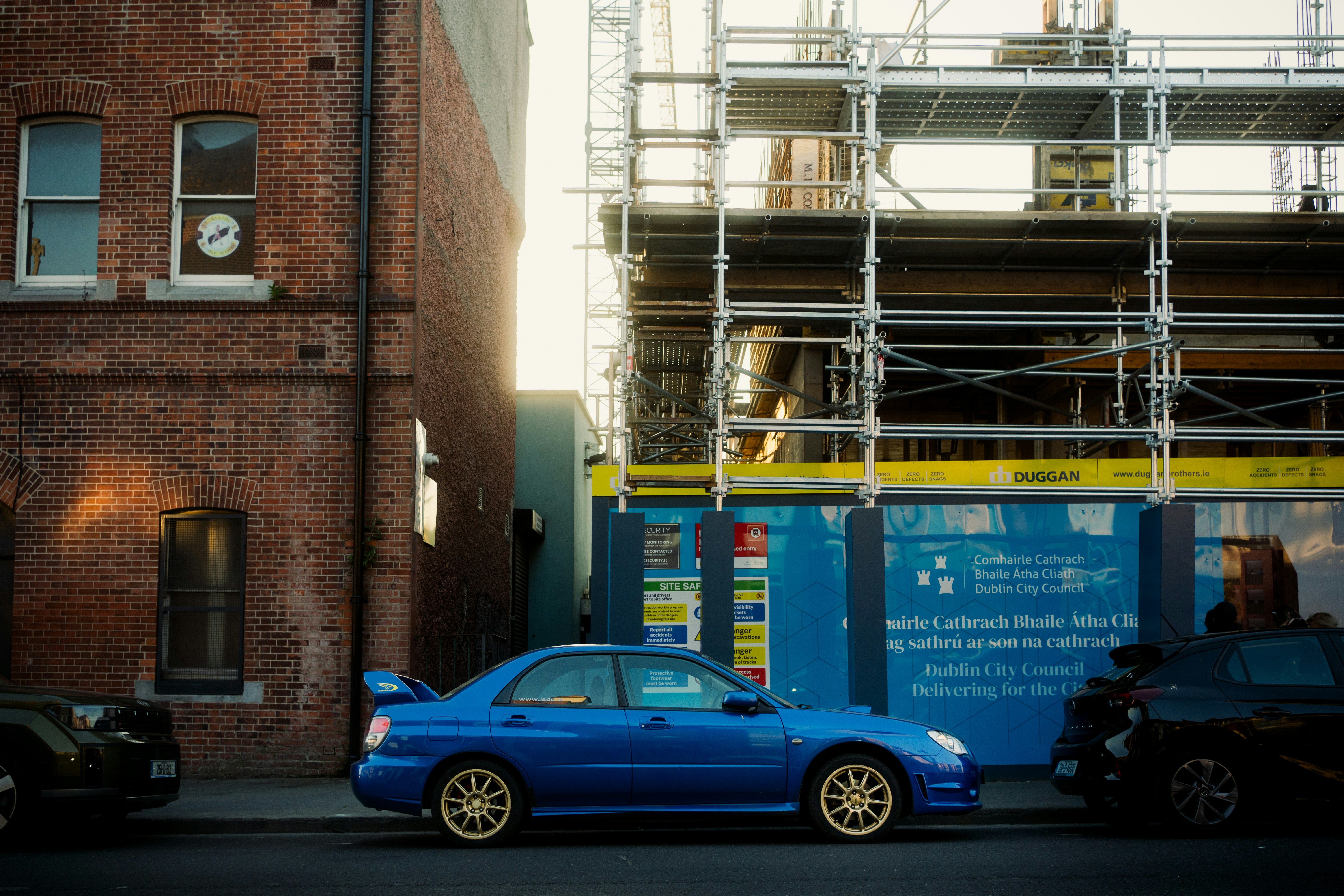 Blue Sedan, Quiet Edge: Urban Scaffold and Brick in Evening Light