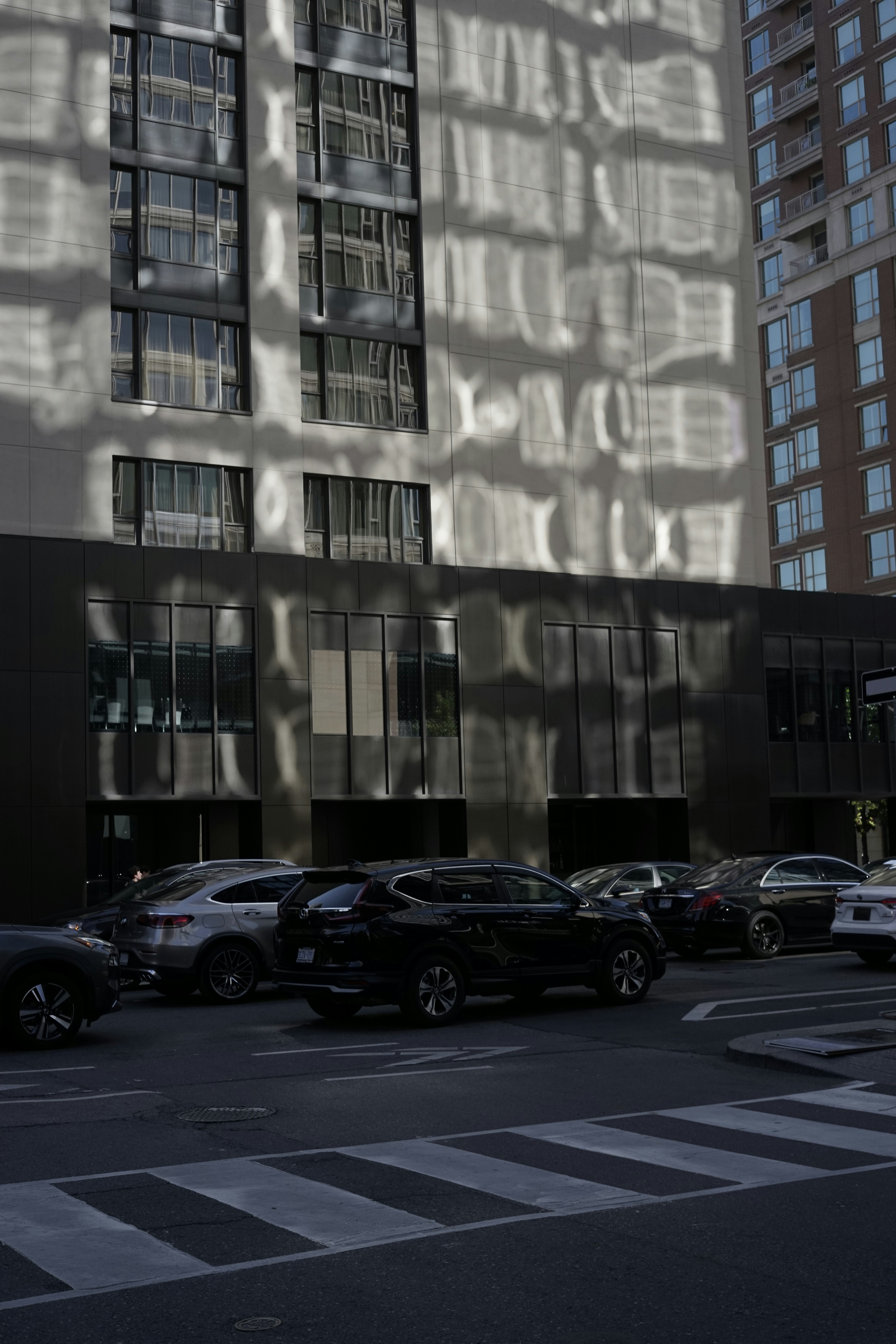 Light patterns cast on a modern building facade, showcasing the interplay of architecture and natural elements. Vehicles line the street below.