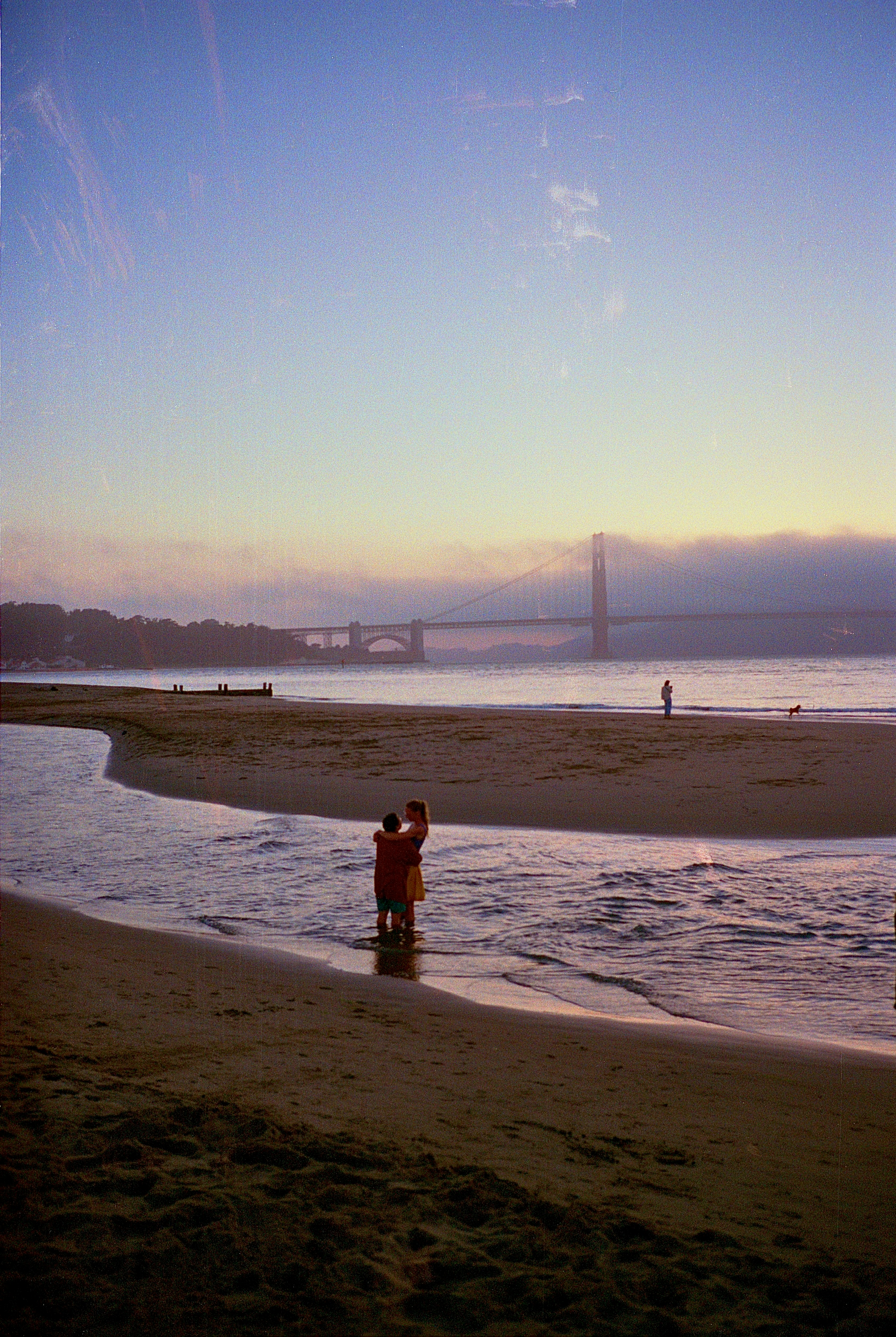 Couple embracing at the water's edge with a bridge silhouetted in the background during twilight. Soft hues reflect the calm atmosphere.