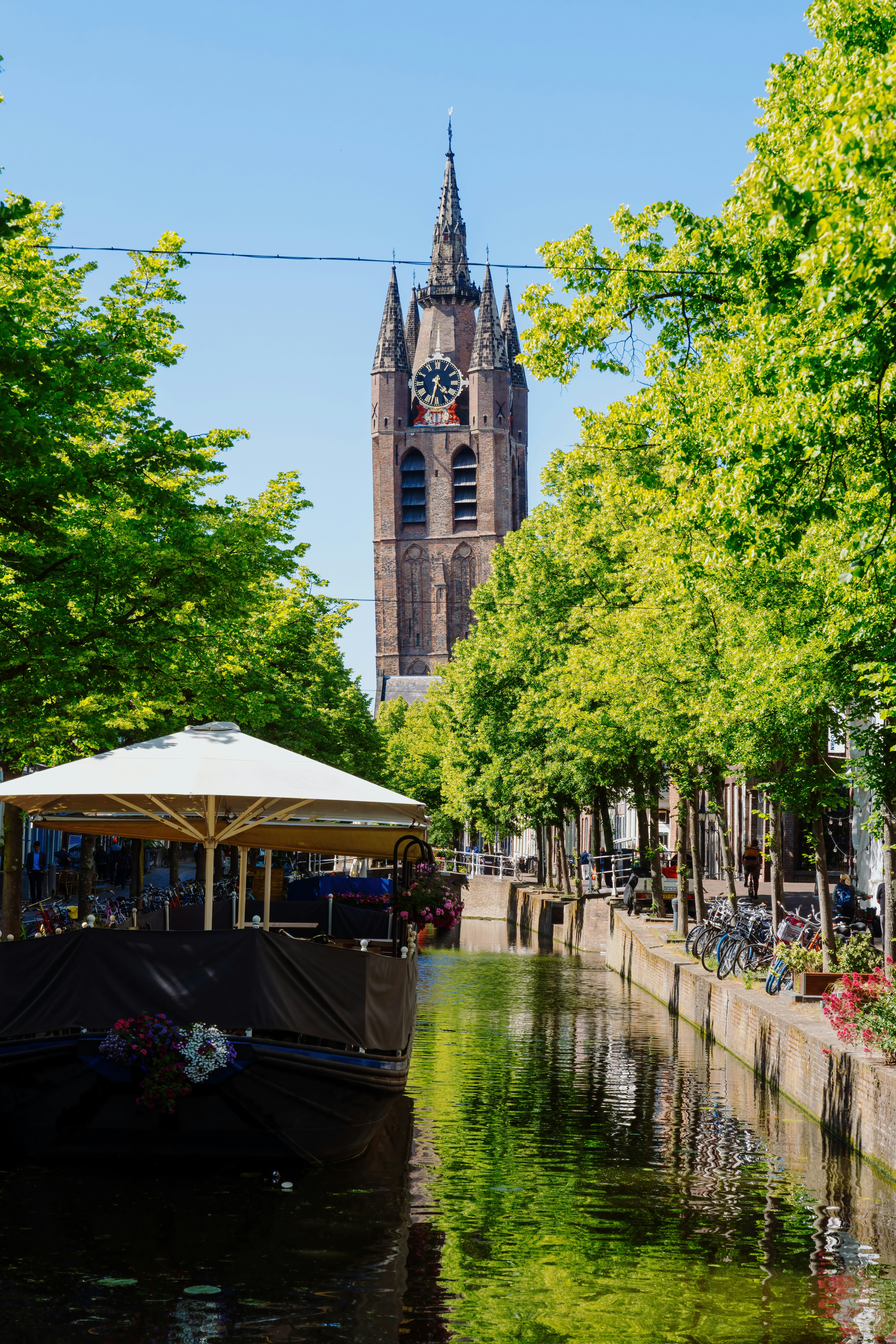 Historic clock tower rising above a serene canal lined with vibrant trees and a boat, creating a peaceful urban landscape.