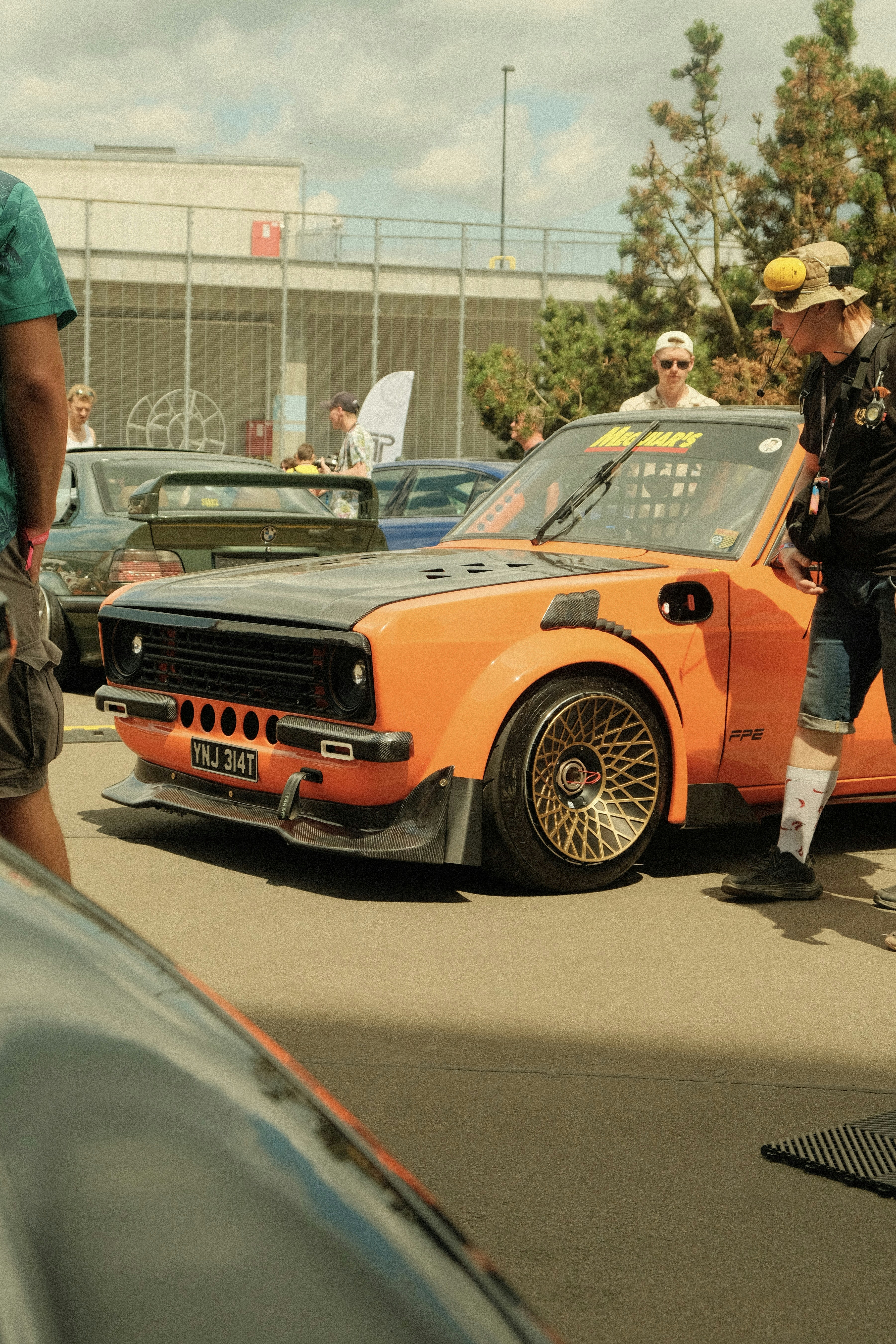 Orange retro racing car with distinctive modifications parked at a car show, surrounded by enthusiasts and classic vehicles.