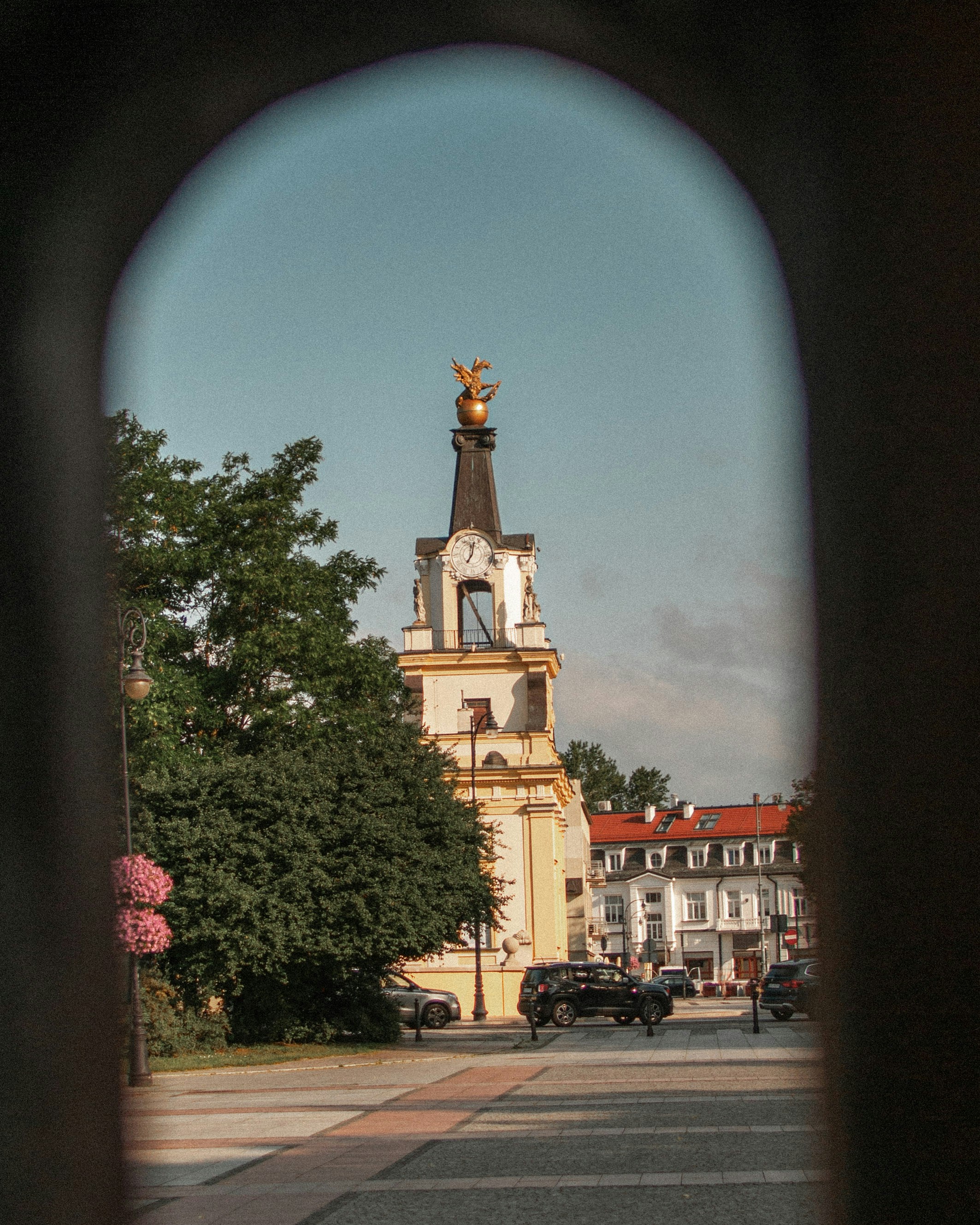 Historic clock tower framed by an archway, surrounded by greenery and urban architecture. The scene captures a blend of nature and city life.
