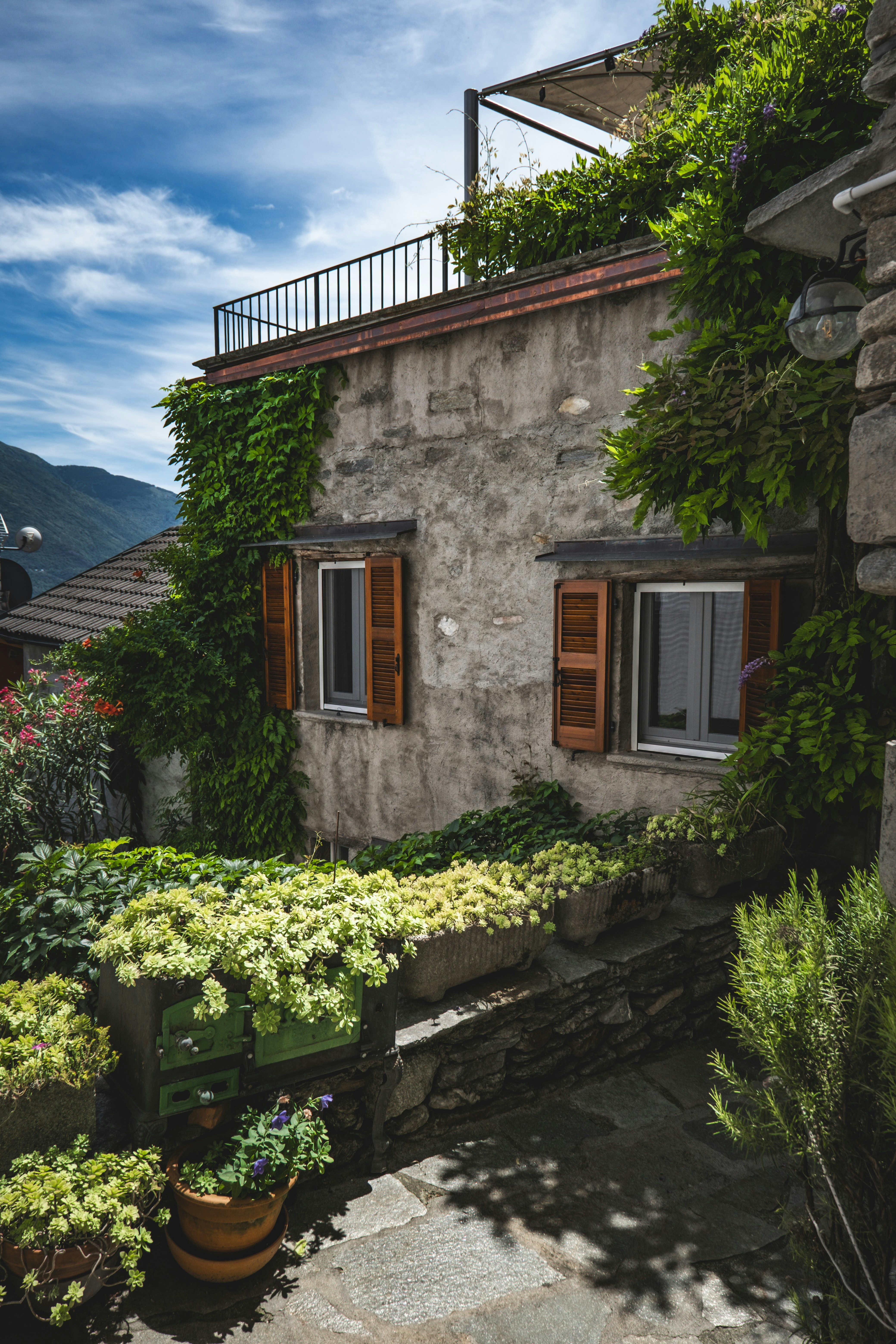 Charming stone building adorned with vibrant greenery and window shutters, set against a backdrop of mountains and a blue sky.