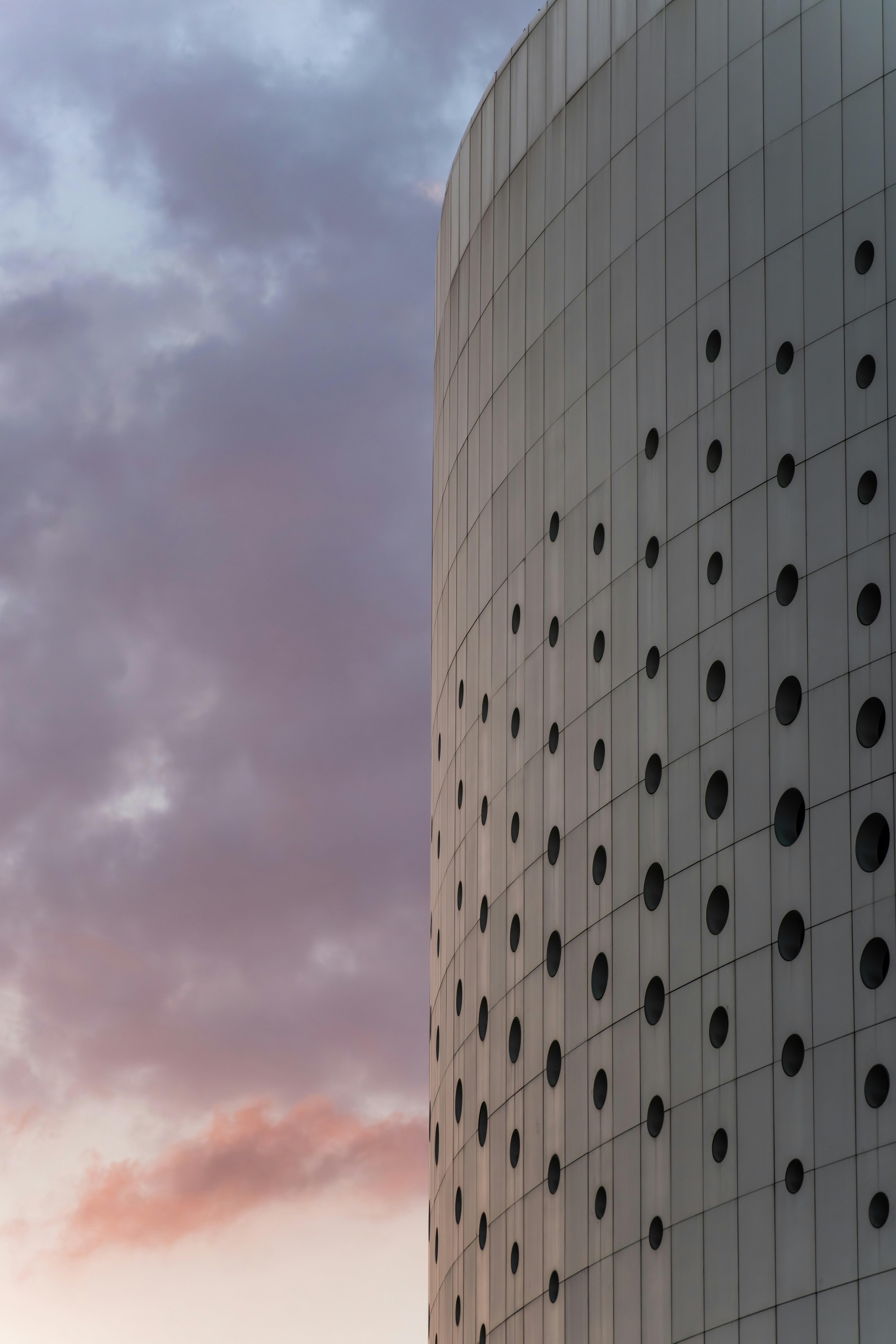 Curved modern building facade adorned with circular perforations, set against a pastel sky at dusk.