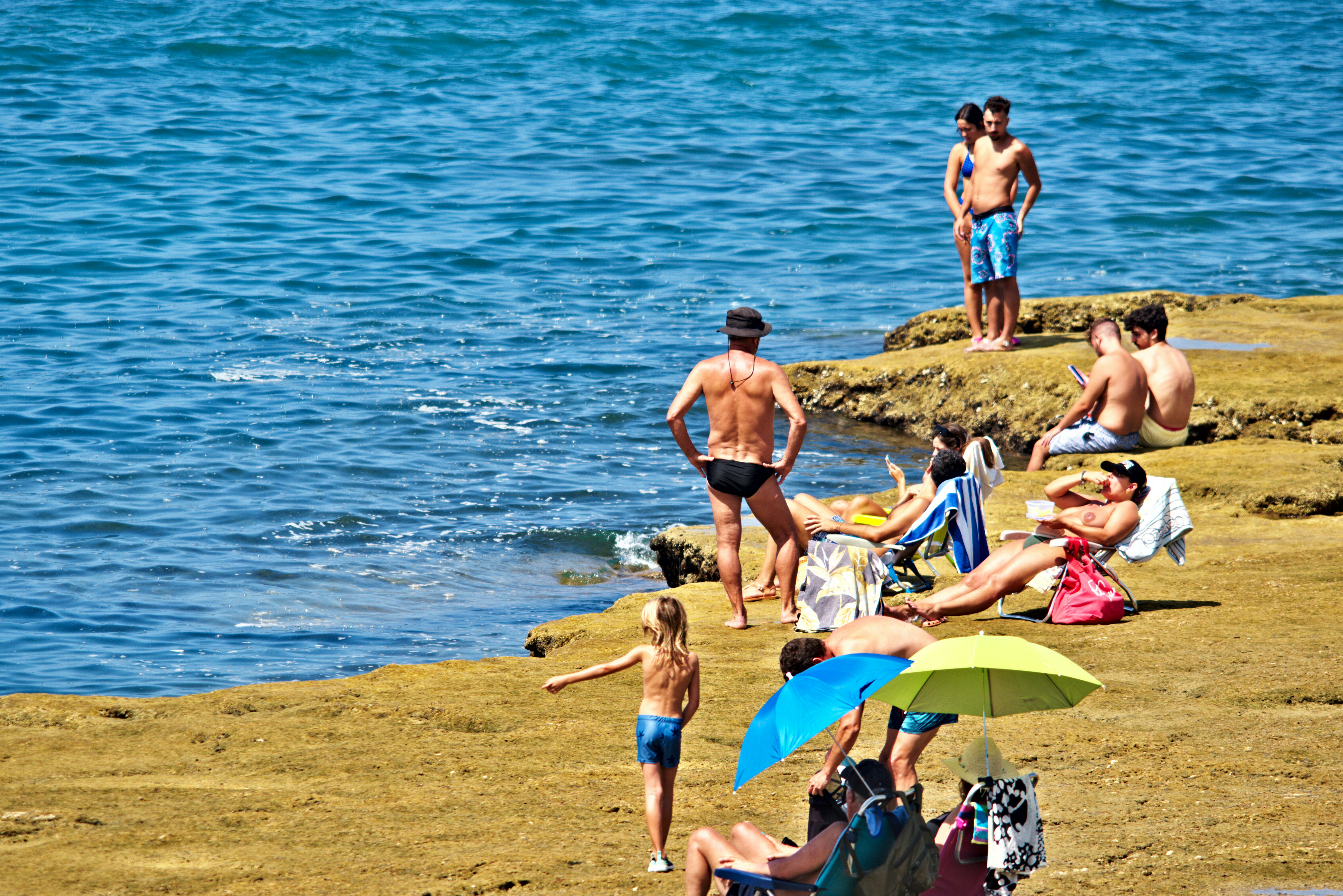 Group of people enjoying a sunny day at the beach, lounging on rocks and playing near the water's edge.