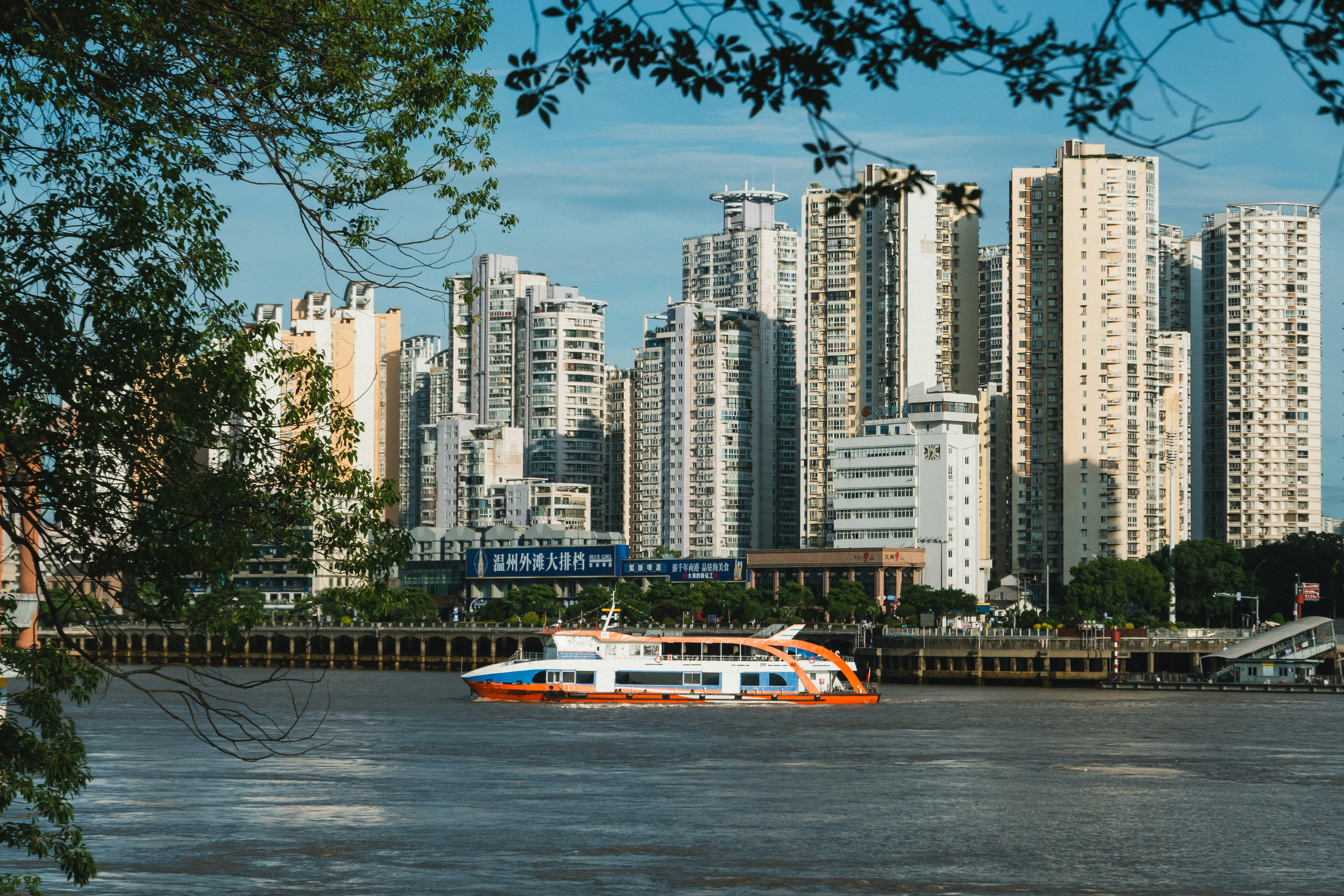 A vibrant ferry glides through the river, framed by a backdrop of towering skyscrapers and lush foliage. The scene captures the blend of nature and urban life.