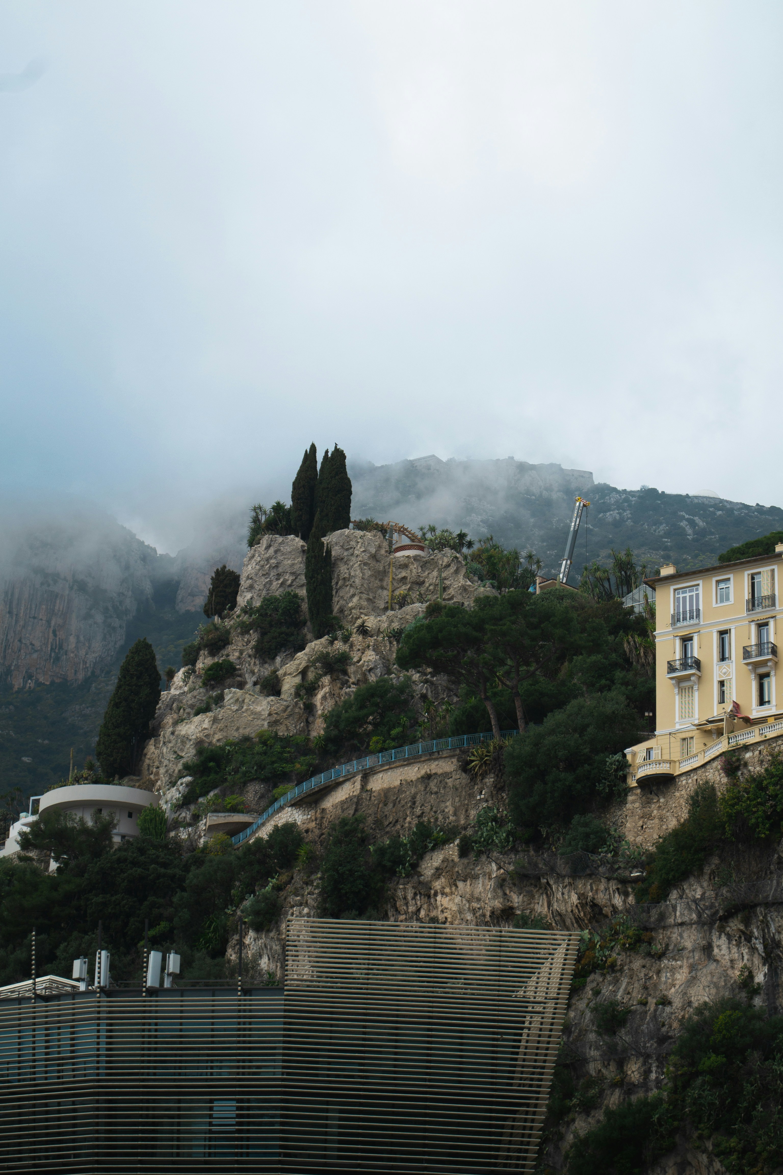 Rocky outcrop adorned with cypress trees, shrouded in mist, overlooking a coastal town. The landscape captures a serene yet dramatic atmosphere.
