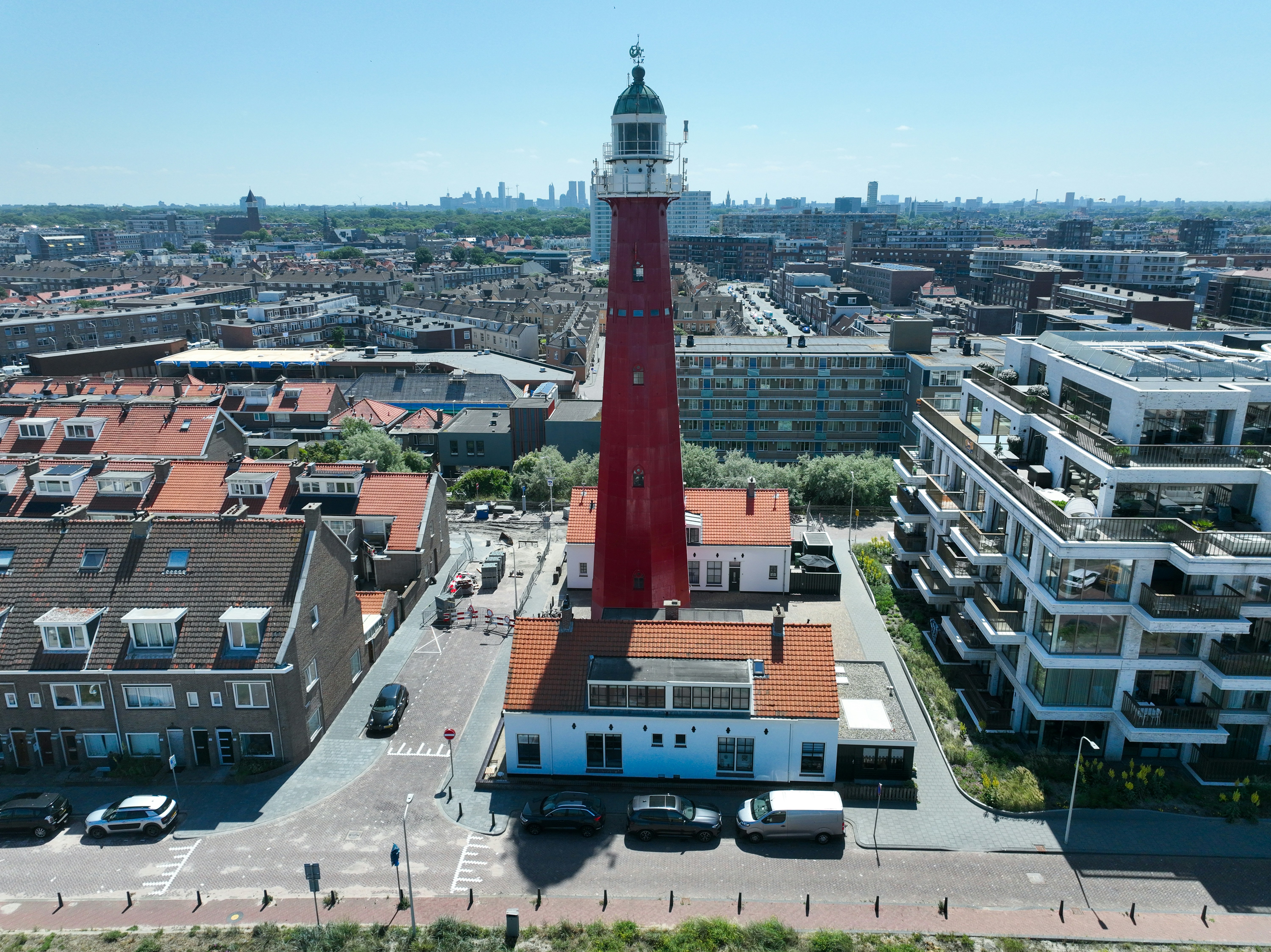 A high-resolution aerial photo of the red lighthouse in Scheveningen, The Hague, taken with a drone. It shows the mix of traditional Dutch homes, modern apartment buildings, and the coastline under clear summer skies. | A tall red lighthouse stands in a sunny town.