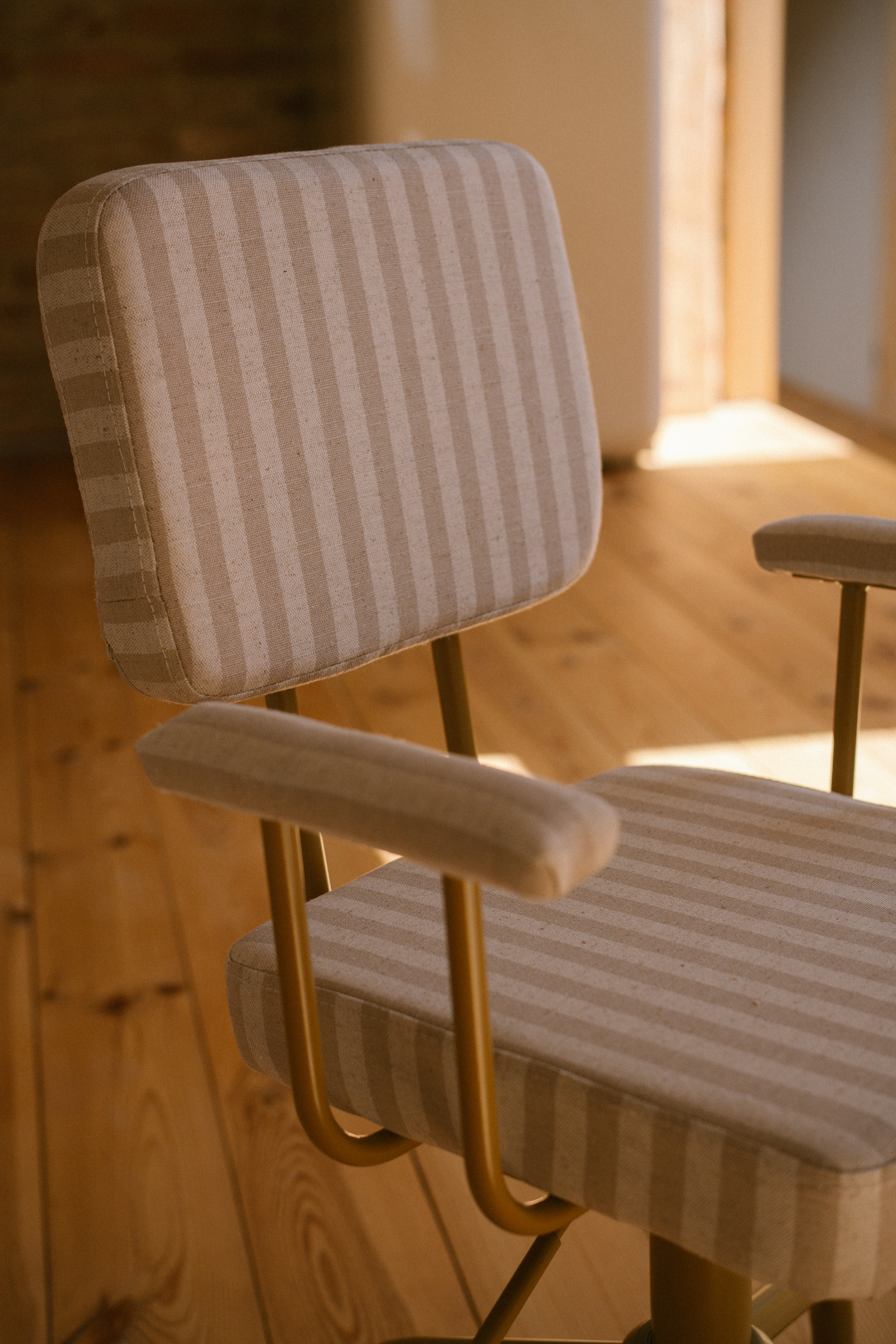 A stylish, striped chair with golden accents positioned in a sunlit room with wooden flooring.
