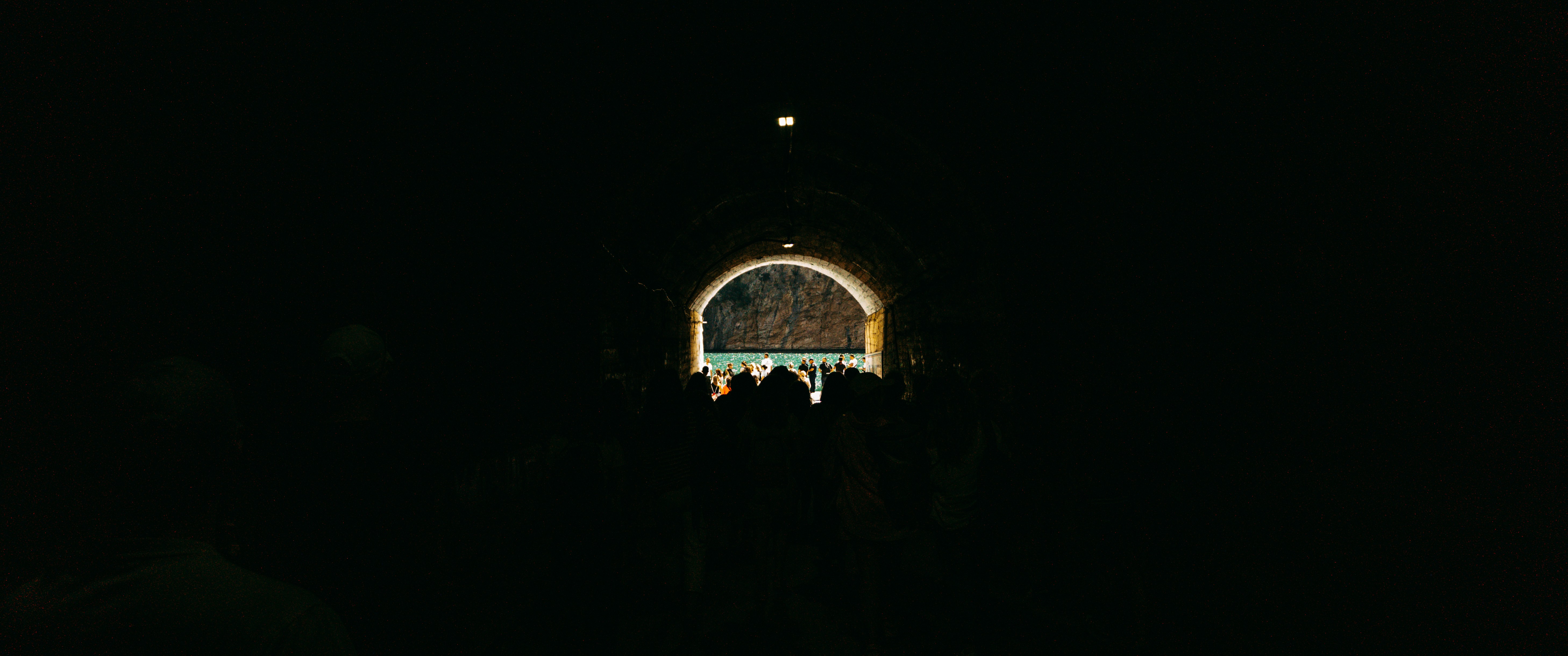 Dimly lit tunnel with a bright archway at the end, surrounded by silhouettes of people. The atmosphere evokes a sense of anticipation and mystery.