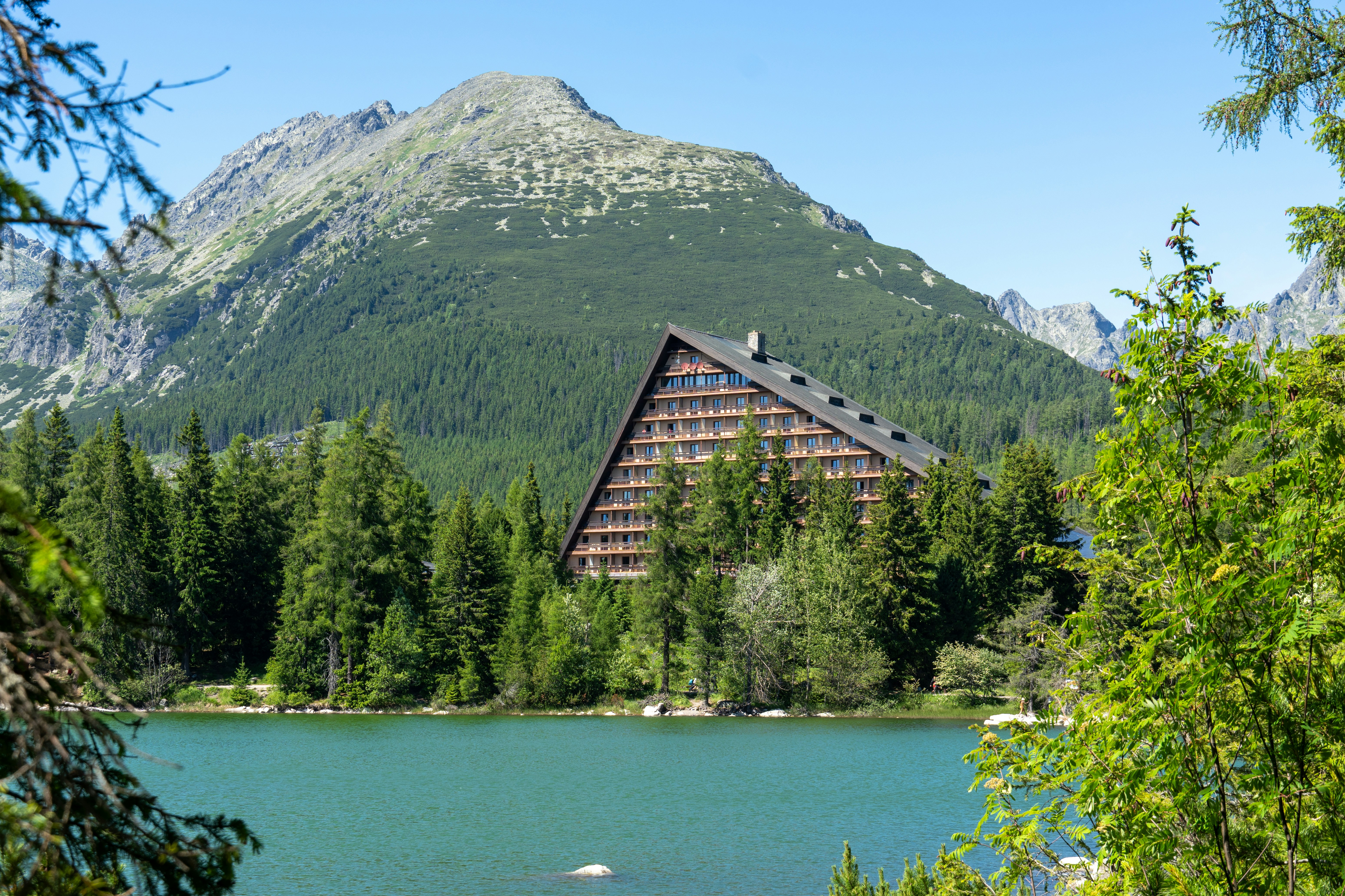 A striking triangular hotel nestled by a serene lake, surrounded by lush forests and majestic mountains in the background.