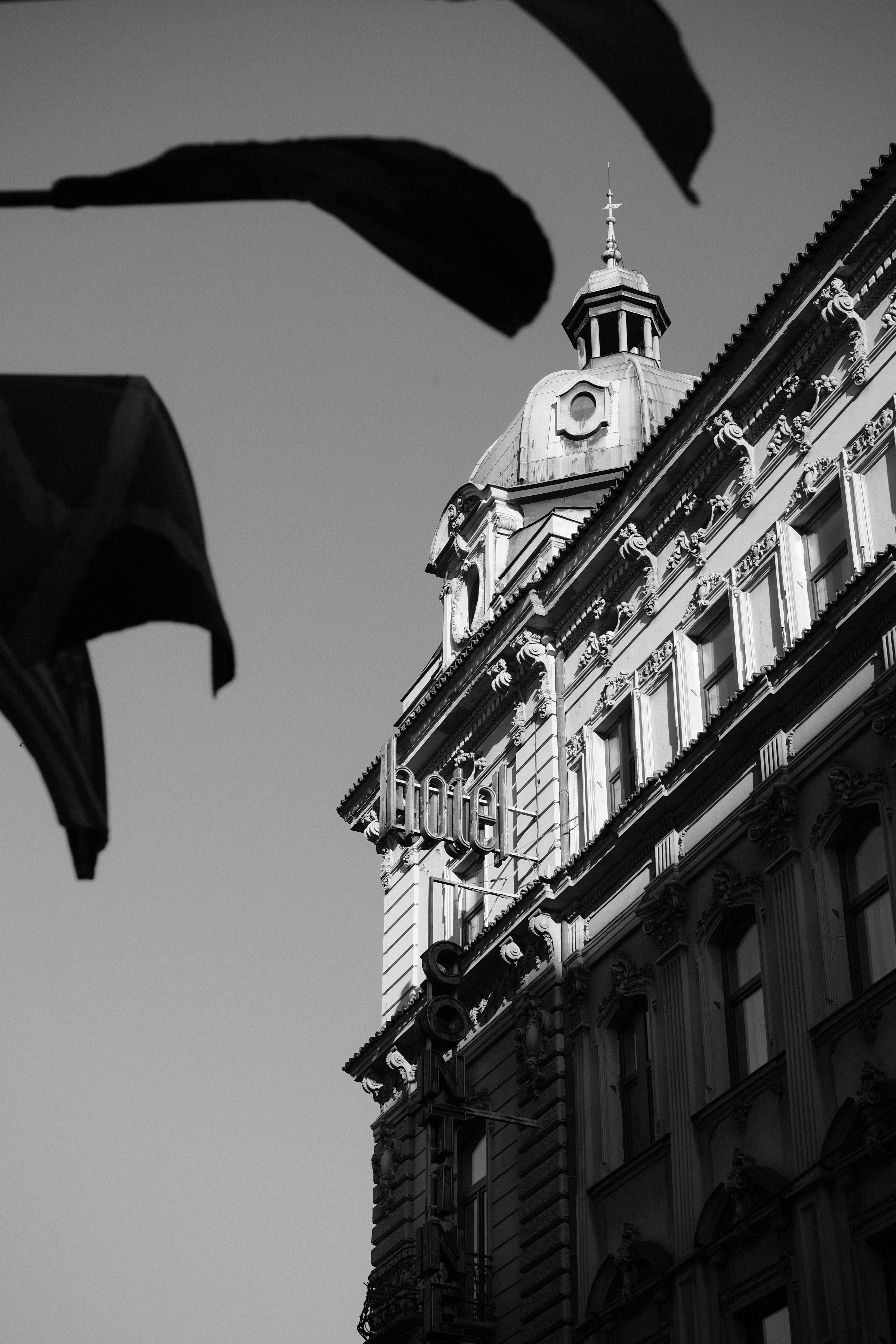 A historic building shows a clock tower and the word hotel on its facade. Flags are partially visible in the foreground against a clear sky. | A hotel building stands proudly against the sky.