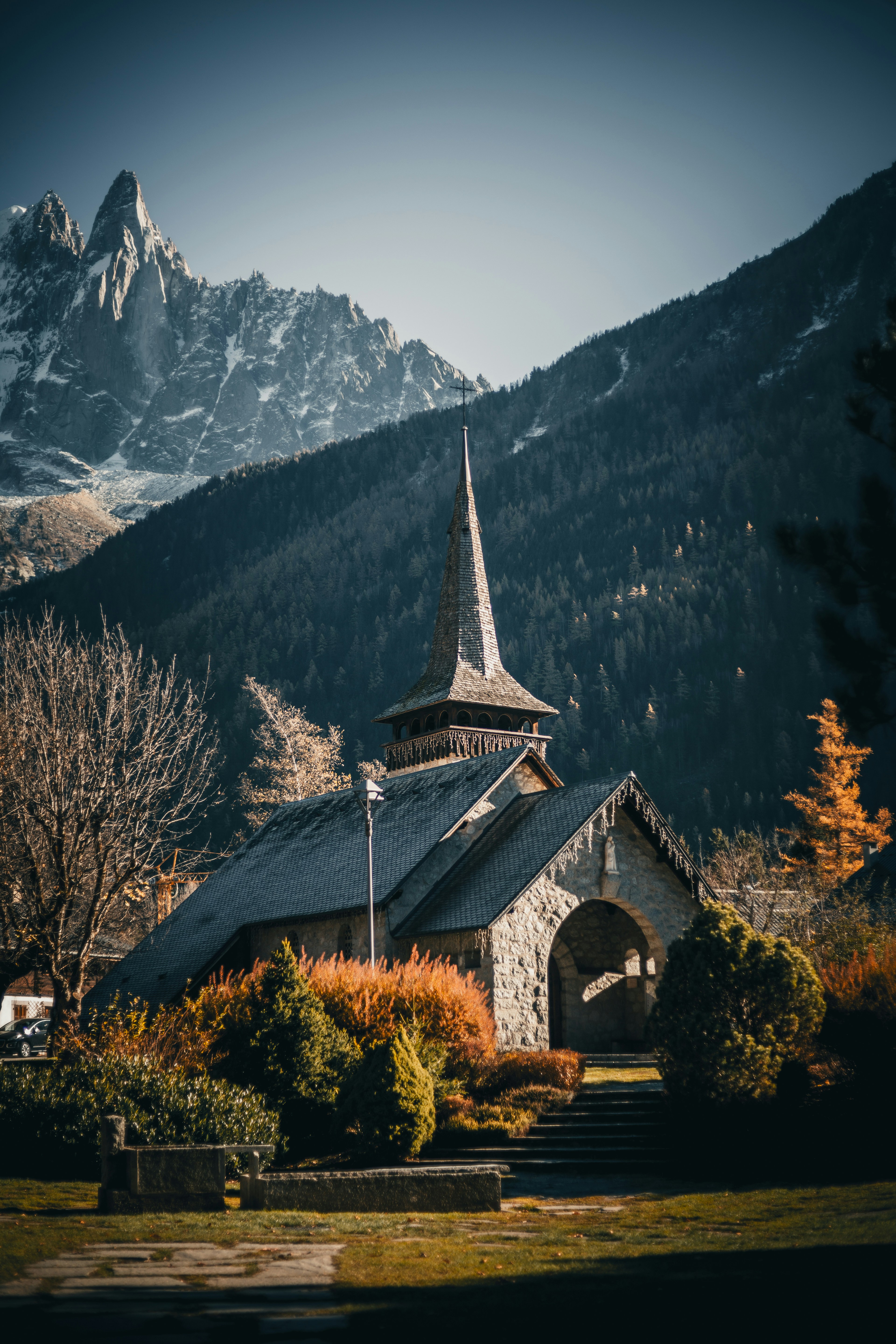 Charming stone chapel nestled in a vibrant landscape, framed by towering mountains and autumn foliage.