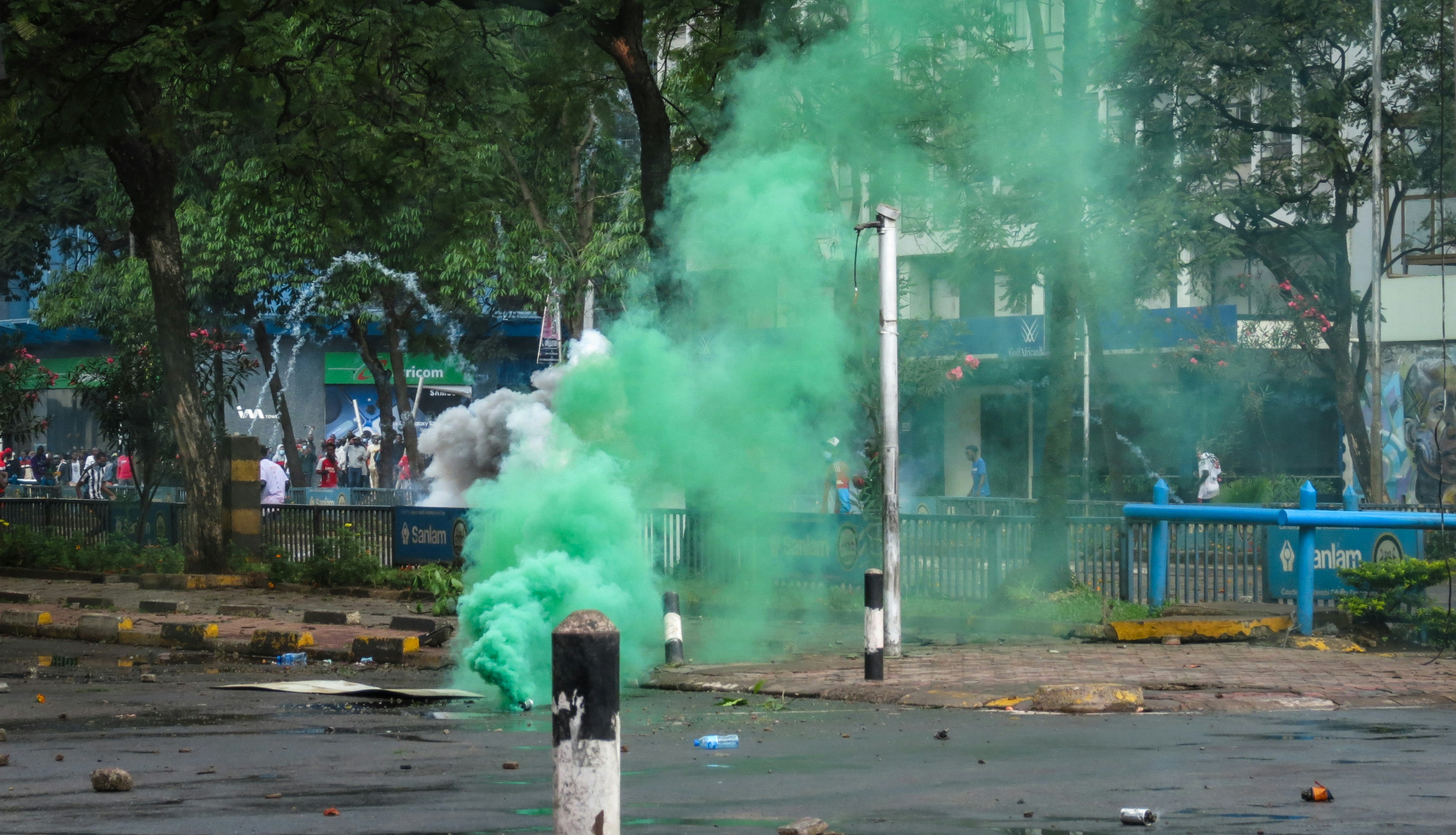 Green smoke billowing from a canister in a street scene, surrounded by debris and a backdrop of urban structures. The atmosphere conveys a sense of unrest.