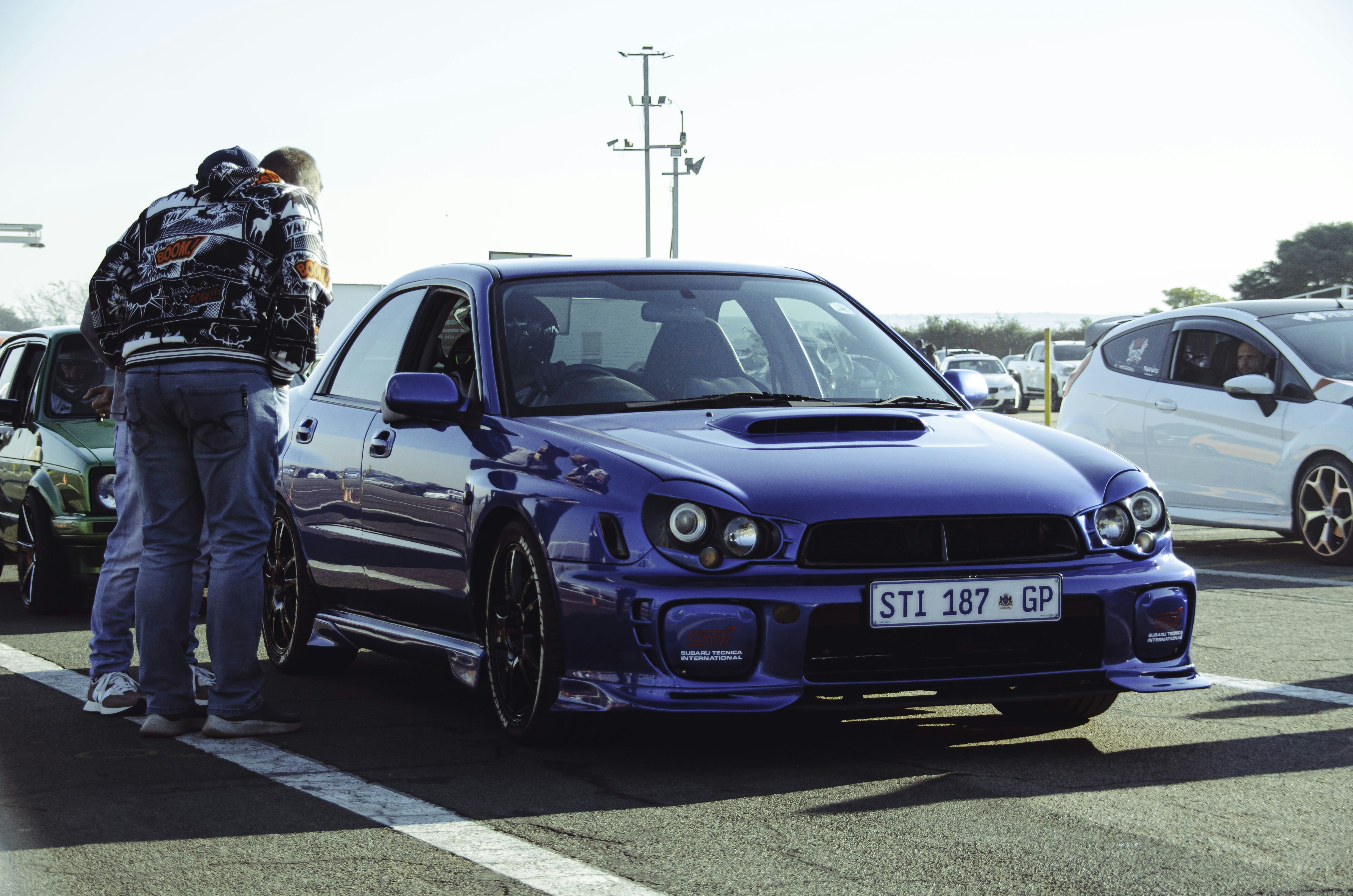 A striking blue sports car parked at a gathering, with an individual inspecting its features. The scene captures the essence of car culture and community.