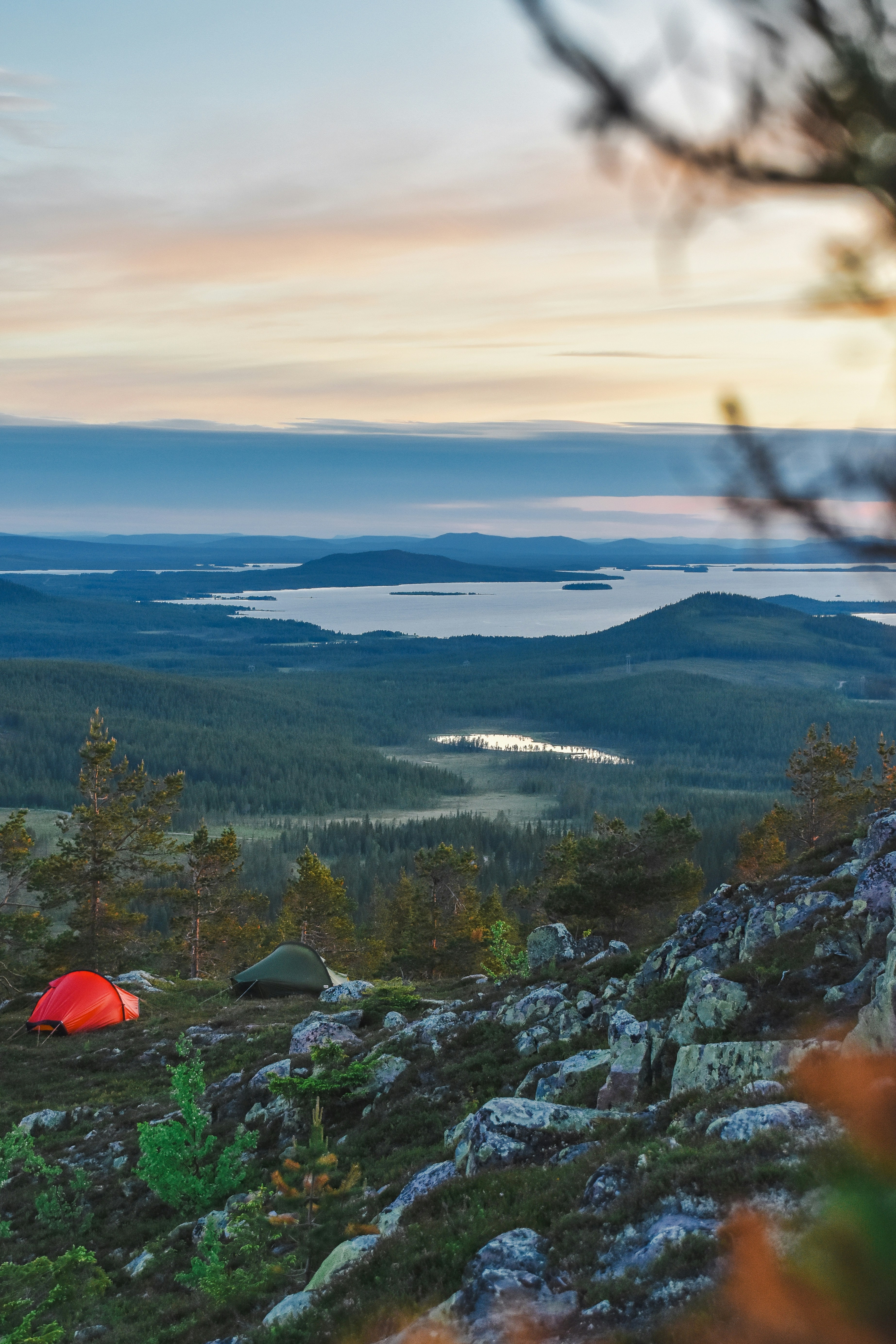 Two tents nestled among rocky terrain, overlooking a vast landscape of lakes and forests under a pastel sky.