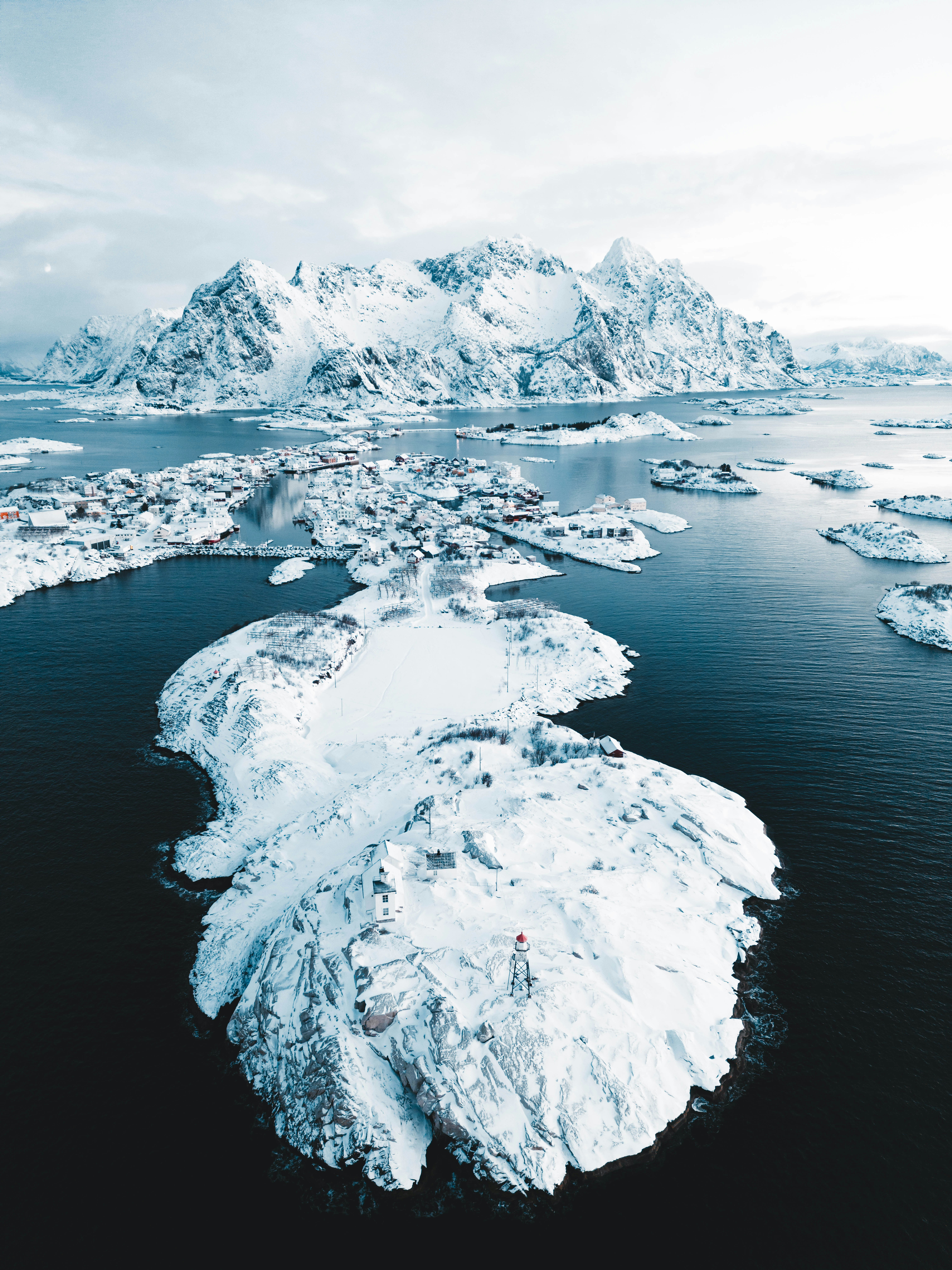 Snow-covered island with a lighthouse surrounded by icy waters and mountains in the background. A quaint village is visible on the shore.