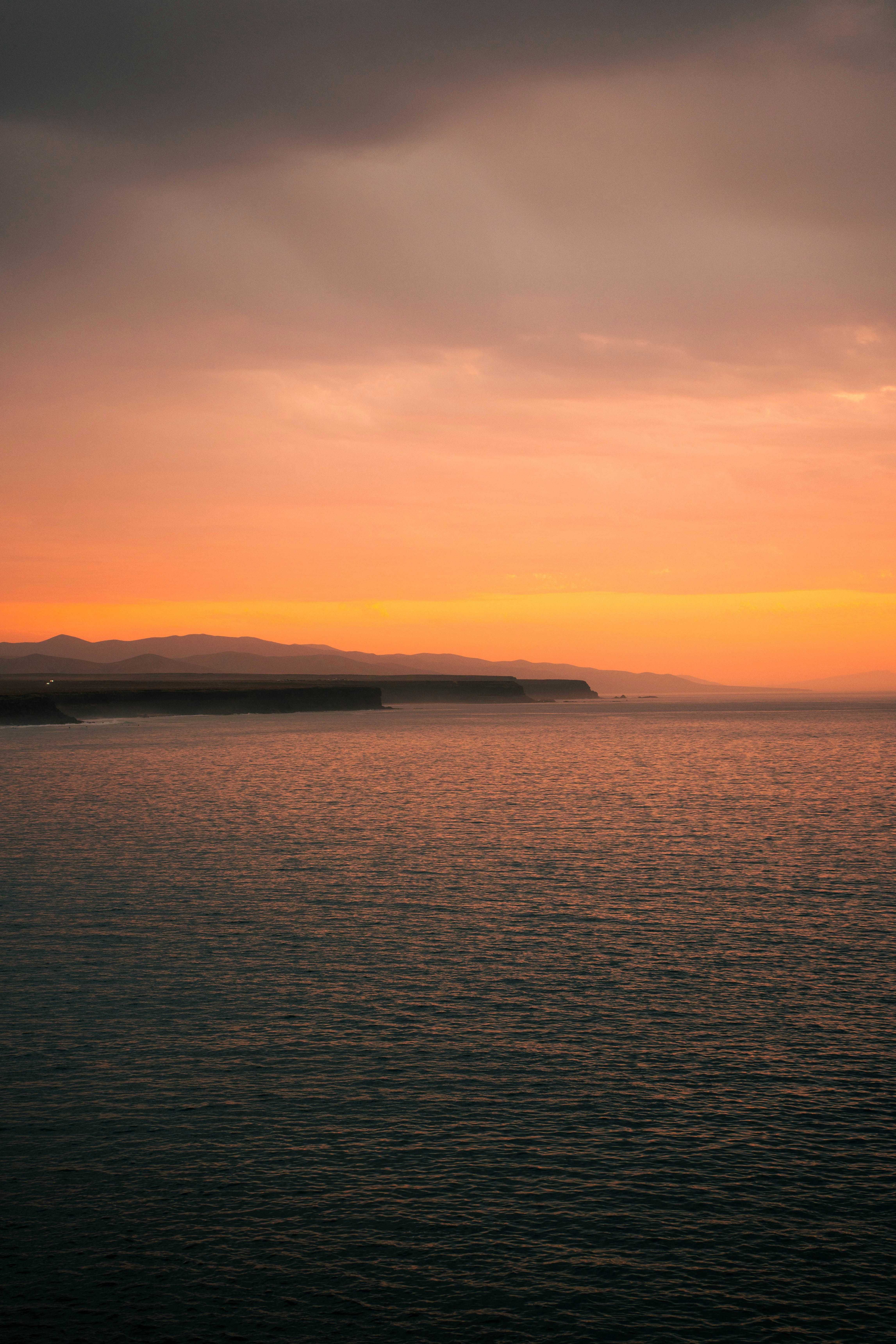 Vibrant sunset hues reflecting on tranquil ocean waters, framed by distant mountains and a dramatic sky.