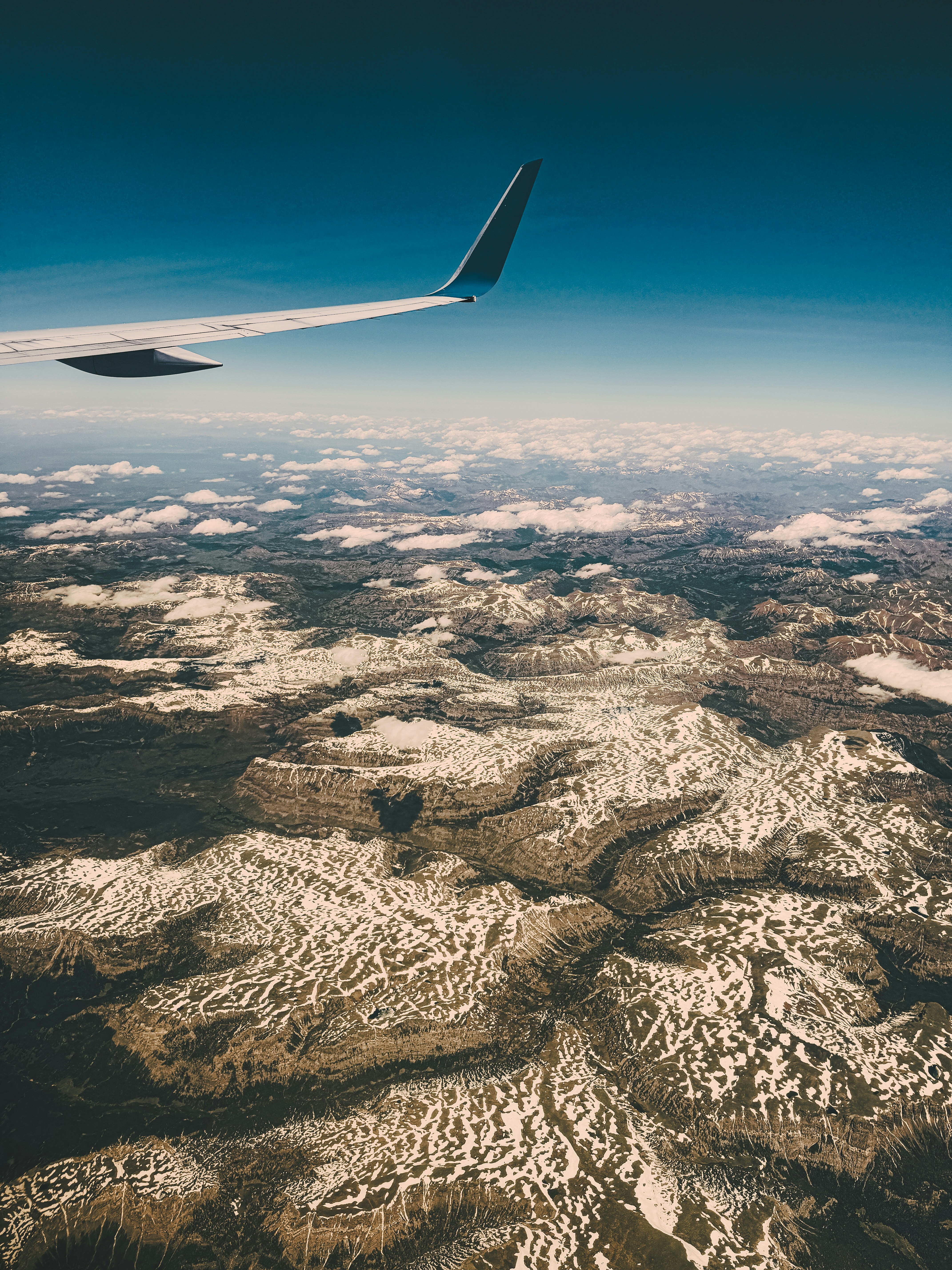 An airplane wing slices across a deep blue sky above a sprawling mountain range dusted with snow. Puffy clouds scatter over the rugged peaks, creating a breathtaking aerial view. | Airplane wing flying above snowy mountains.