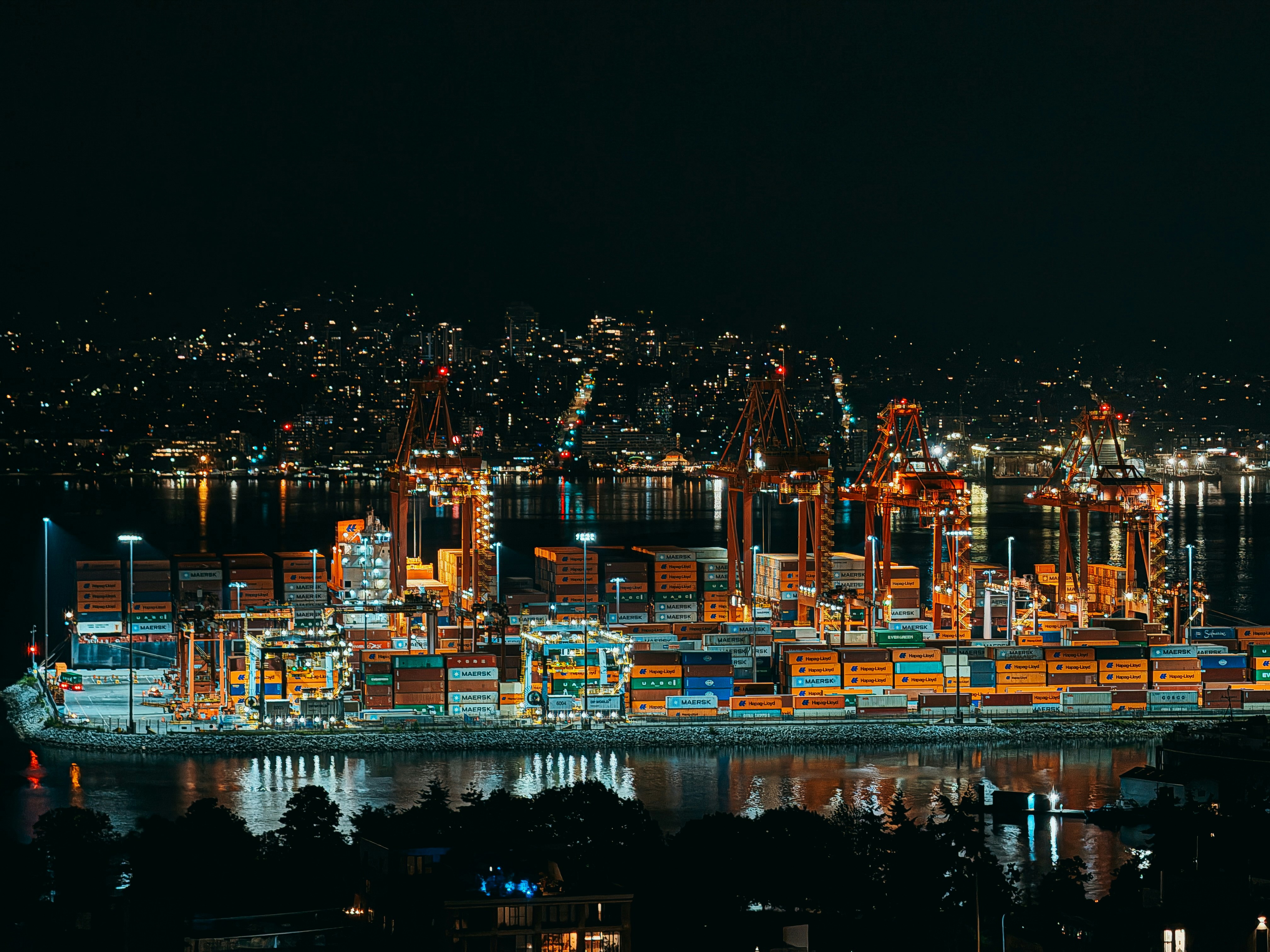 Brightly lit shipping cranes and colorful containers create a striking industrial scene at a busy port, with city lights twinkling in the background across the water. The nighttime setting emphasizes the vibrant energy and scale of global trade. | Nighttime view of a brightly lit cargo port.