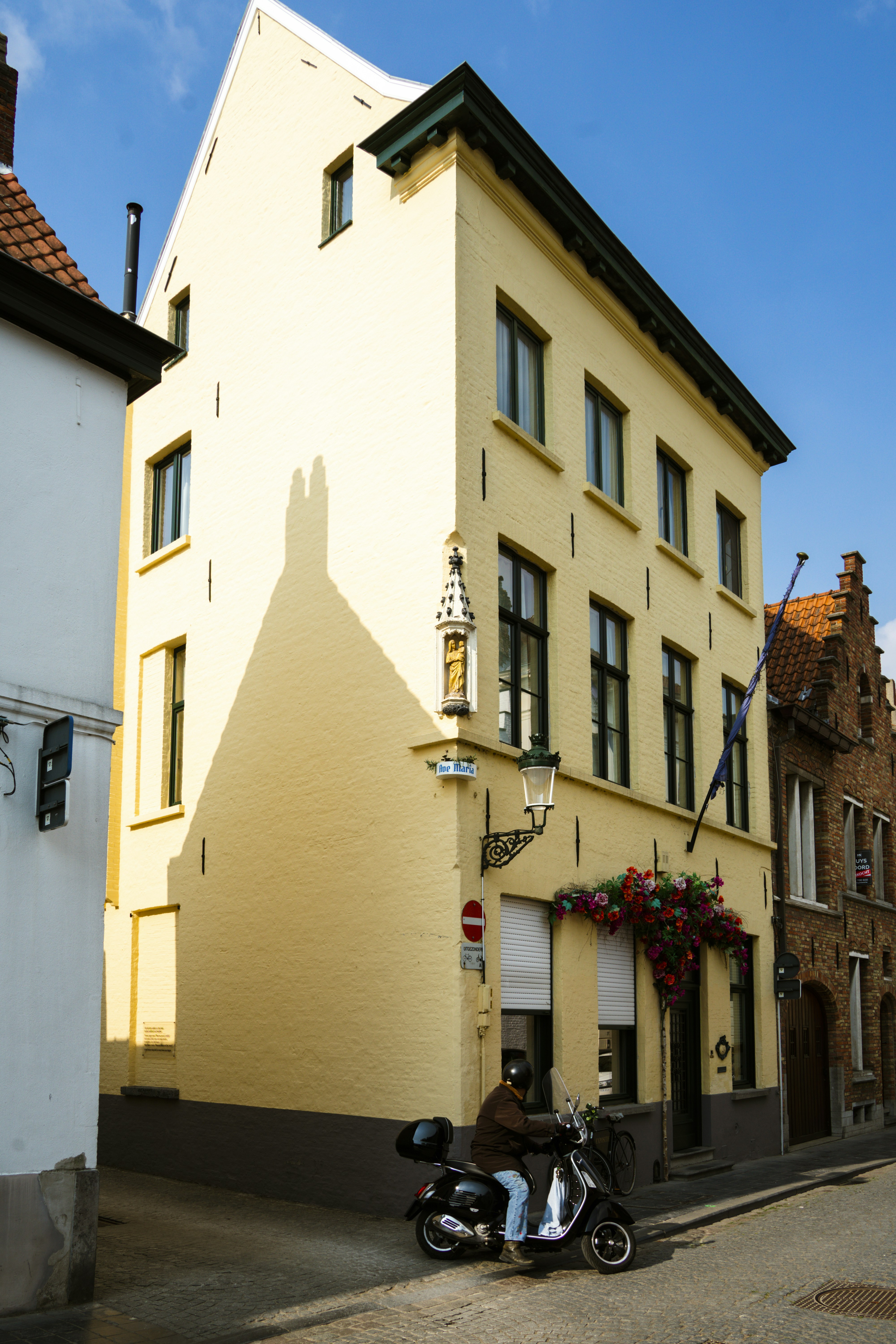 A quaint yellow building adorned with vibrant flowers and a statue casts a shadow, while a rider on a scooter pauses nearby.
