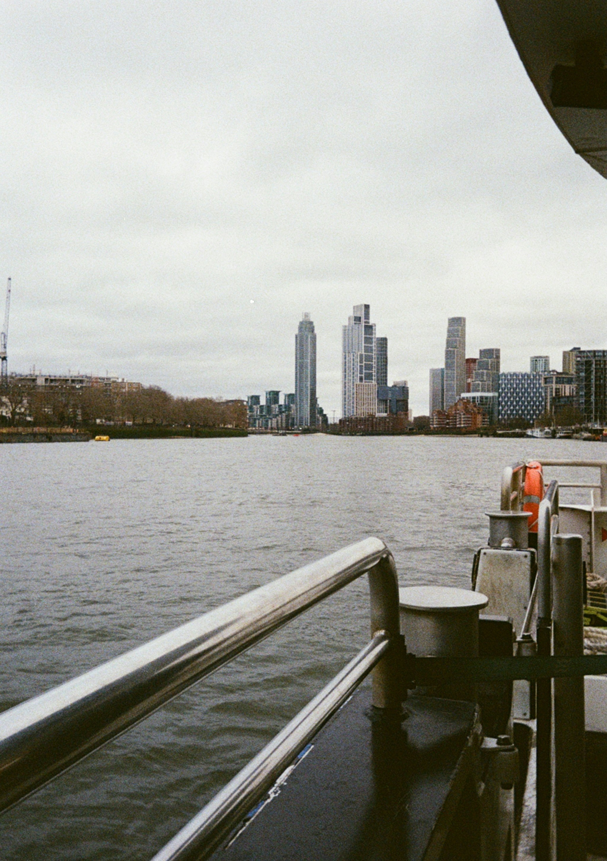 City skyline along a wide river viewed from a ship's railing; foreground includes metal rail and deck machinery, mid-river water, distant buildings under an overcast sky.