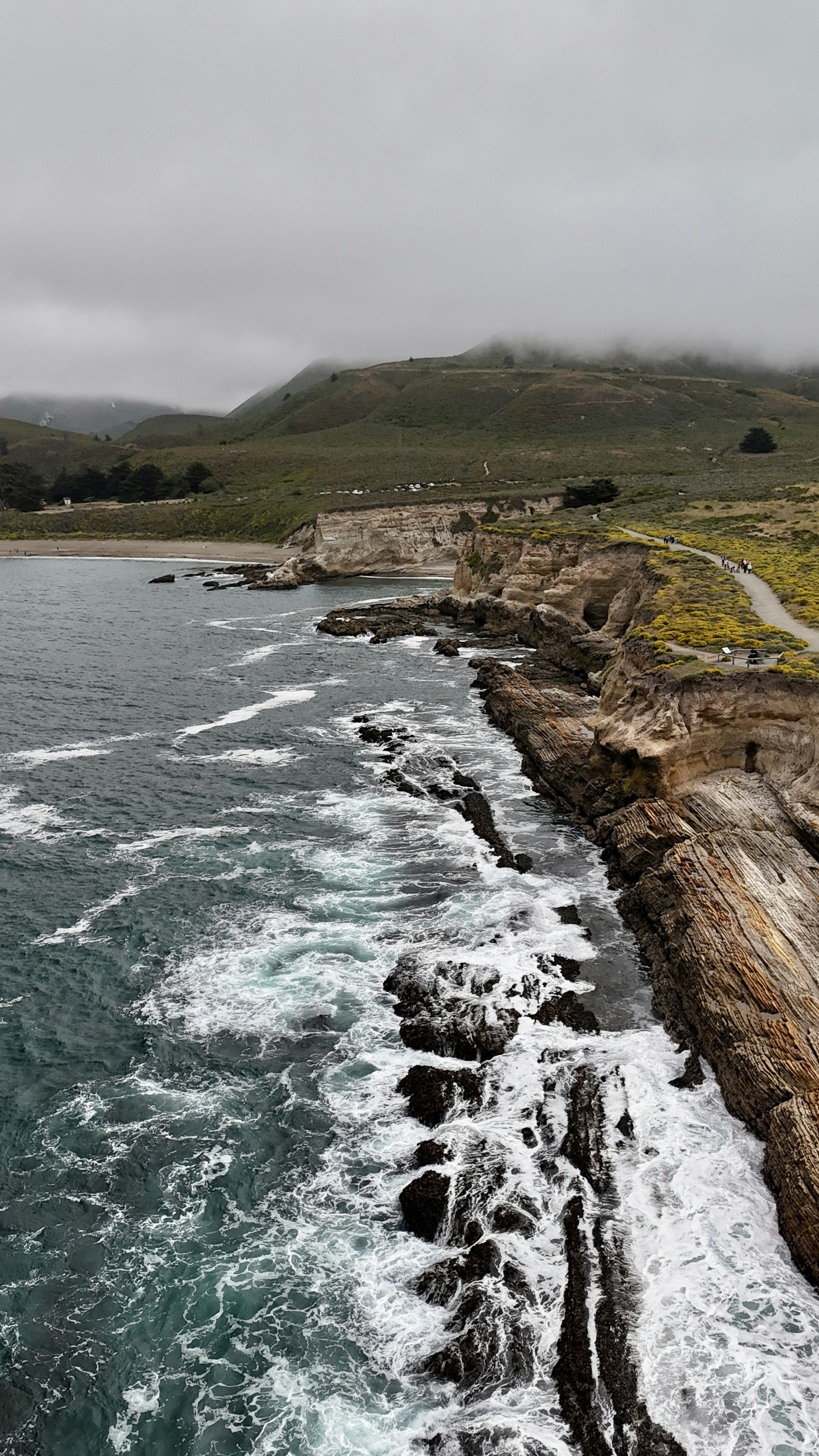 Waves break against layered rock ledges along a rugged cliffside trail, while mist clings to the hills in the background under a heavy sky. The vertical composition captures the raw energy of the coastline at Montaña de Oro State Park. | Waves crash against a rocky coastline on a cloudy day.