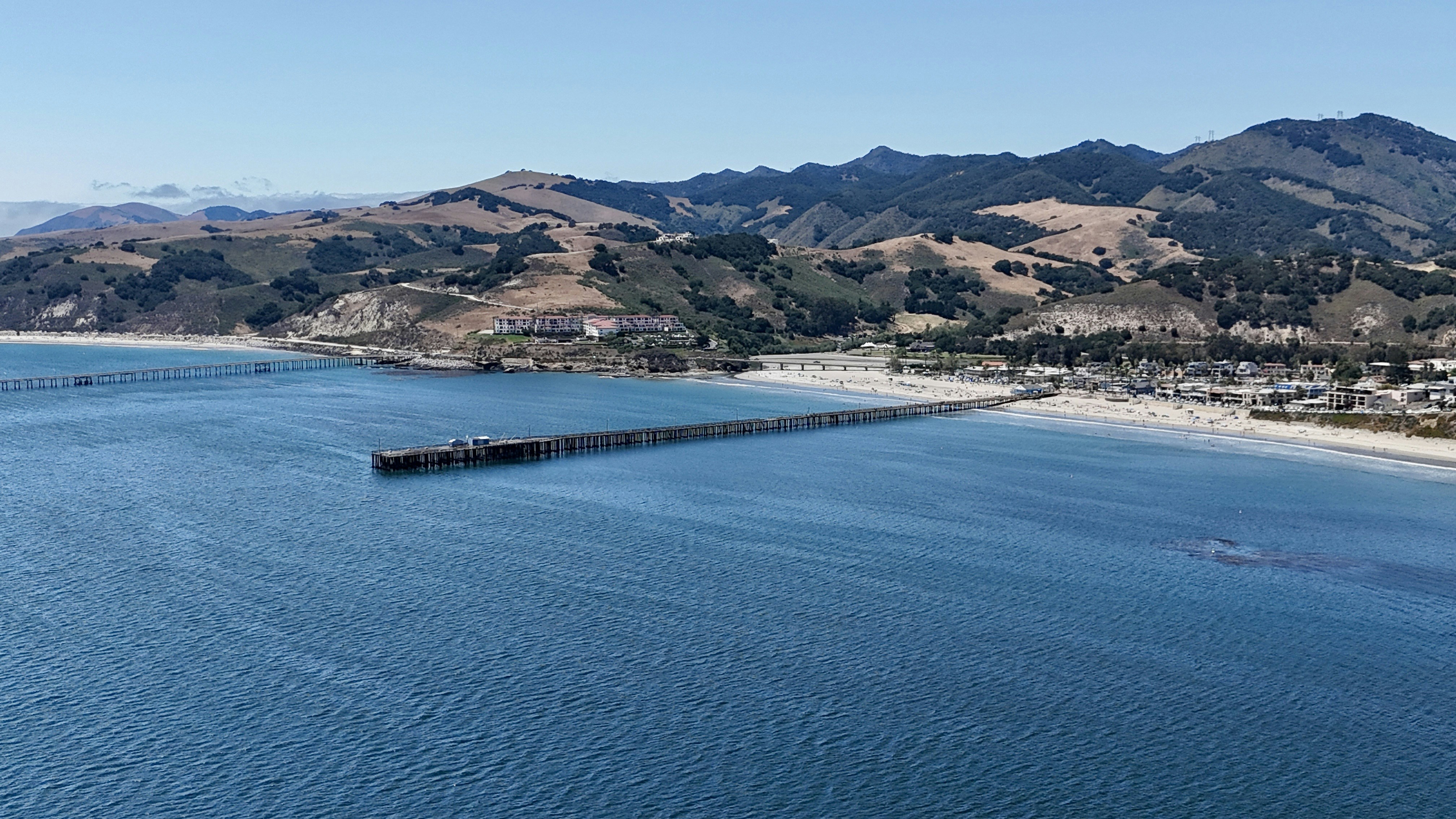 Aerial view of a long pier extending into the tranquil waters, framed by rolling hills and a serene beach. Ideal for capturing the harmony between land and sea.