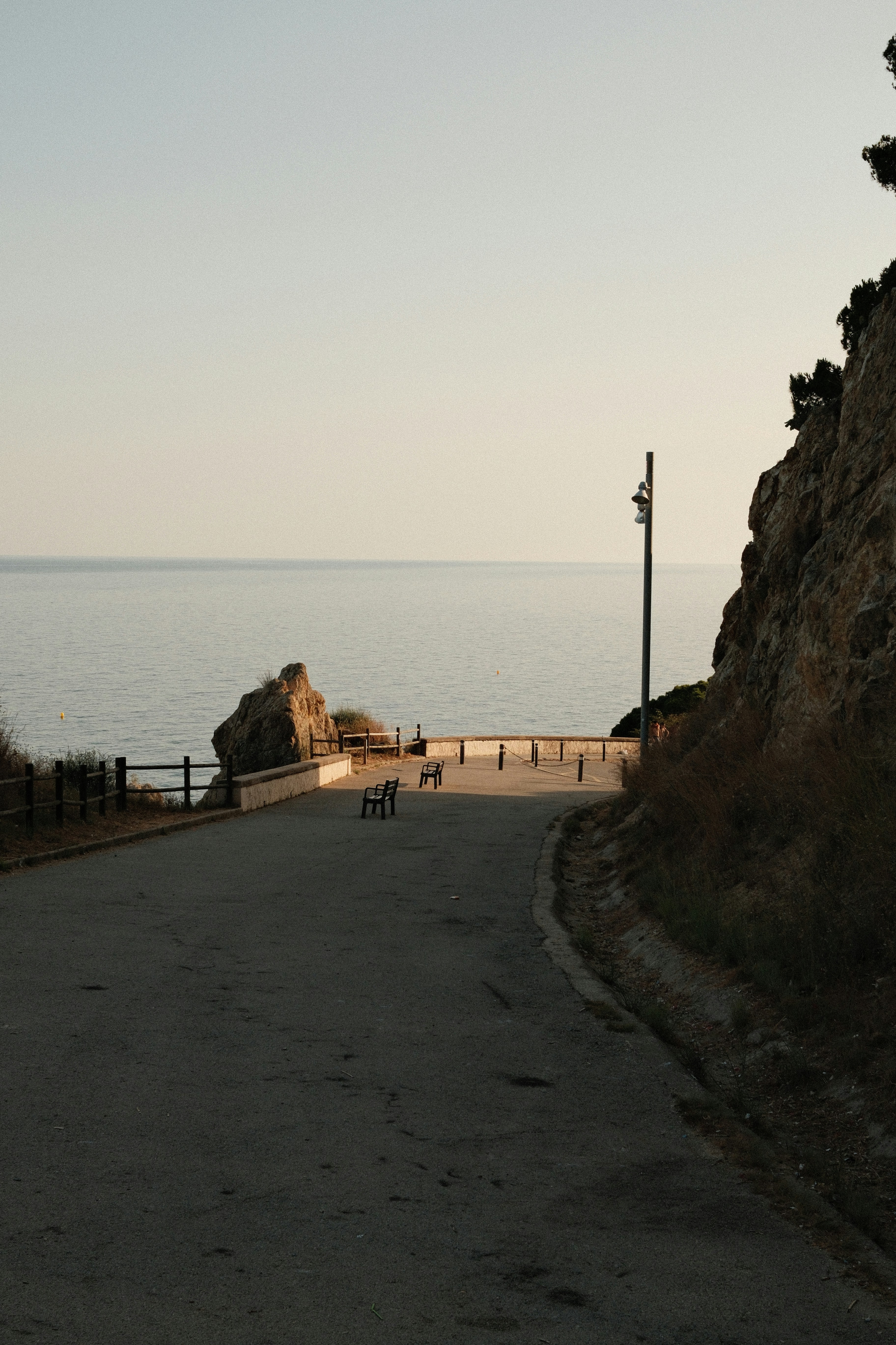 Quiet coastal road leading to the sea, framed by rocky cliffs and sparse vegetation. Two chairs sit invitingly along the path.
