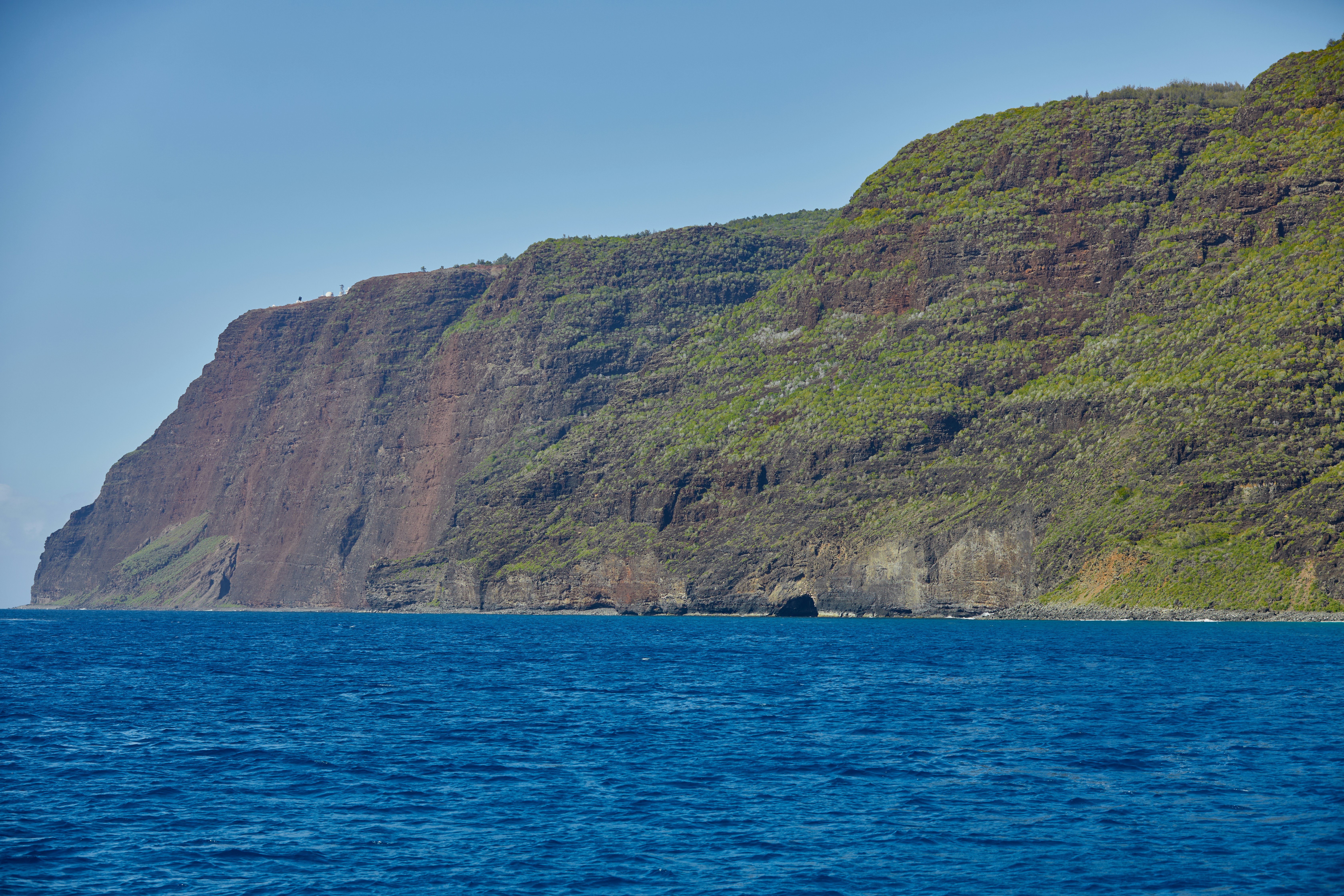 Lush green cliffs rise dramatically from the deep blue ocean, showcasing the rugged beauty of the coastline. The interplay of light and shadow highlights the geological features.
