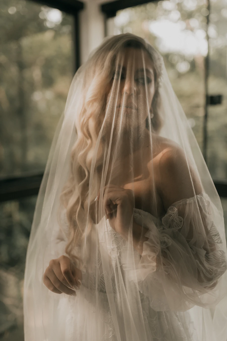 A bride stands behind a sheer veil, her face softly obscured by the fabric. Warm natural light and blurred greenery frame the intimate portrait.